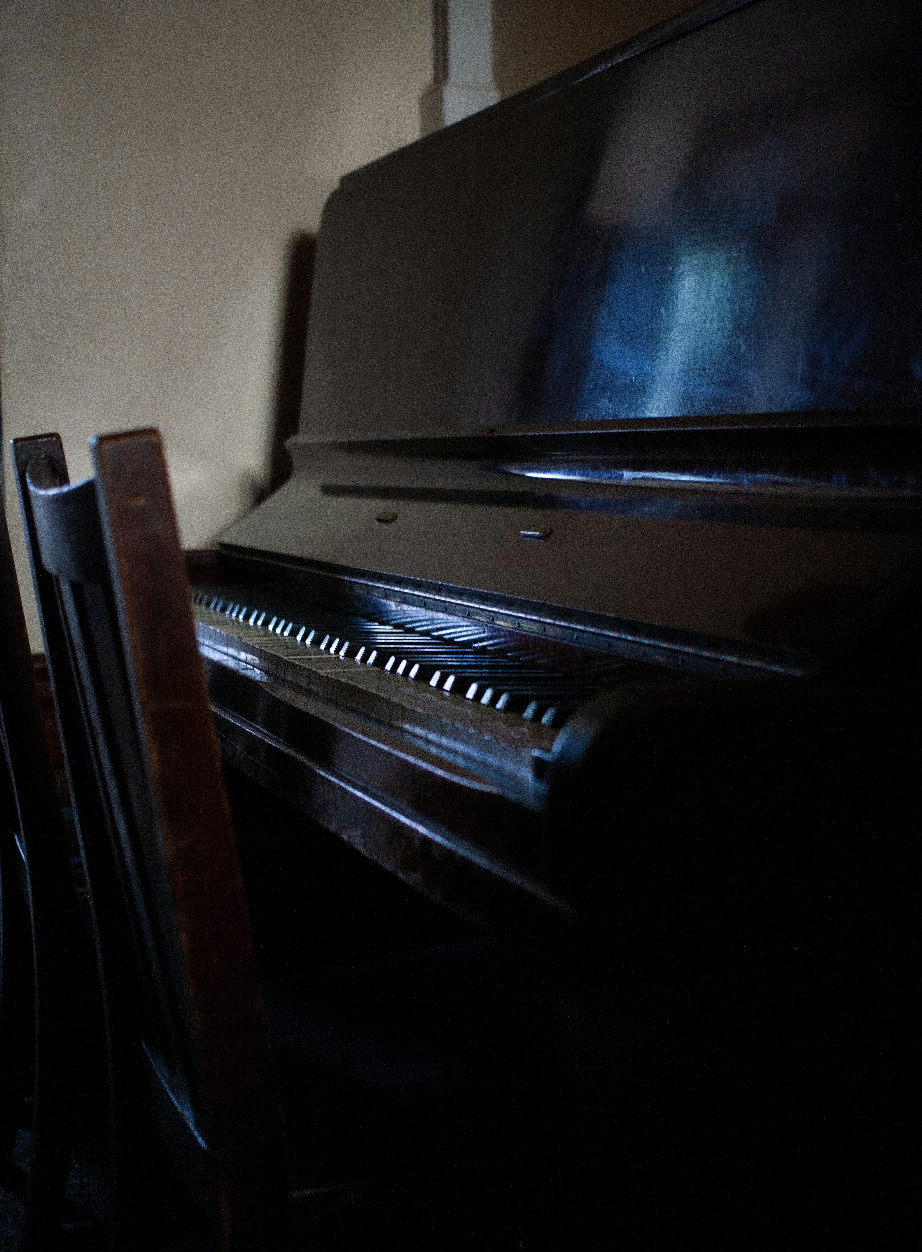 Dimly lit photograph of a glossy upright piano with dark wooden chairs nearby. Blue reflections glide across the lid and keys, highlighting the instrument's sheen.