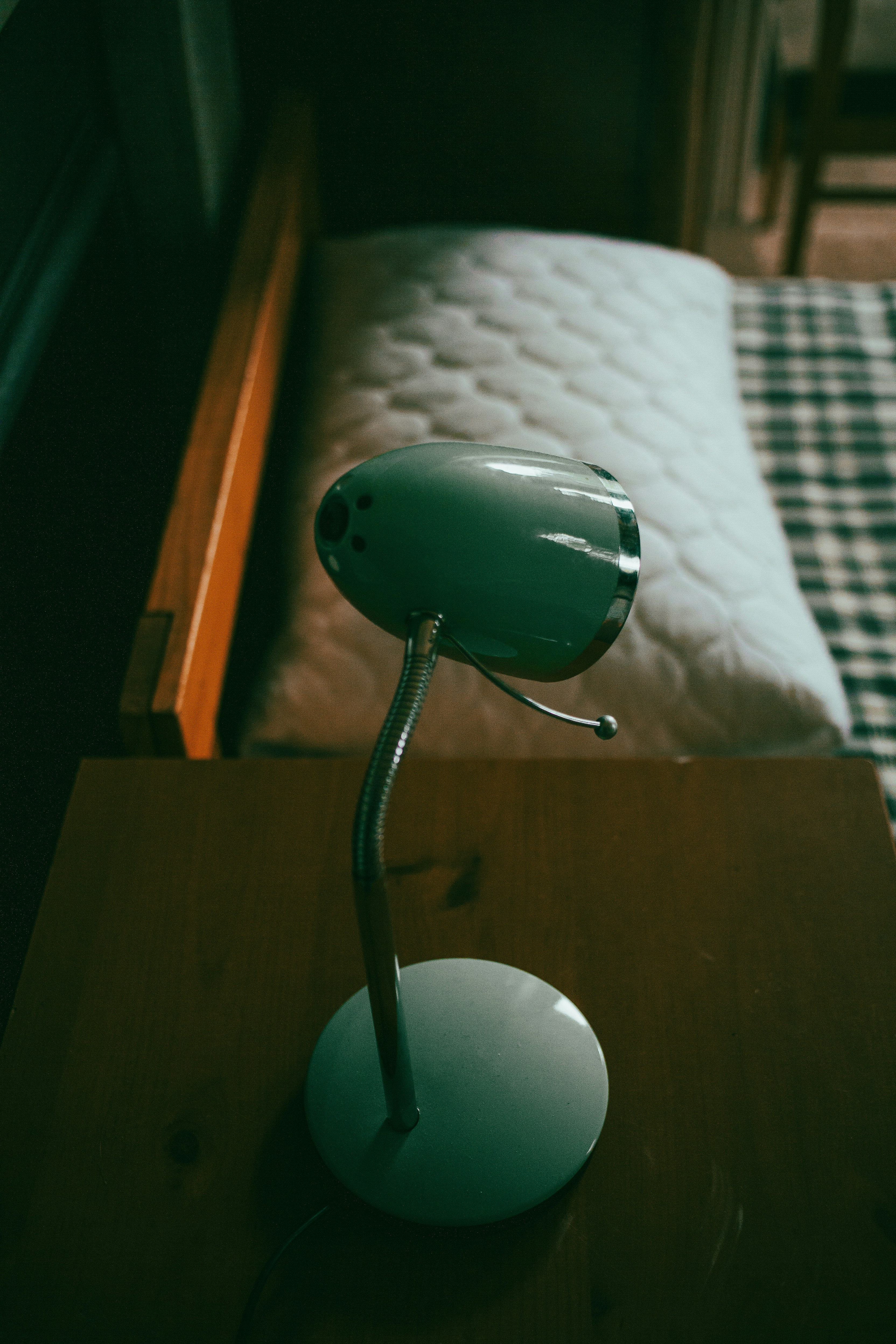 Green gooseneck desk lamp on a wooden table in a dimly lit room, with a cushioned headboard in the background.