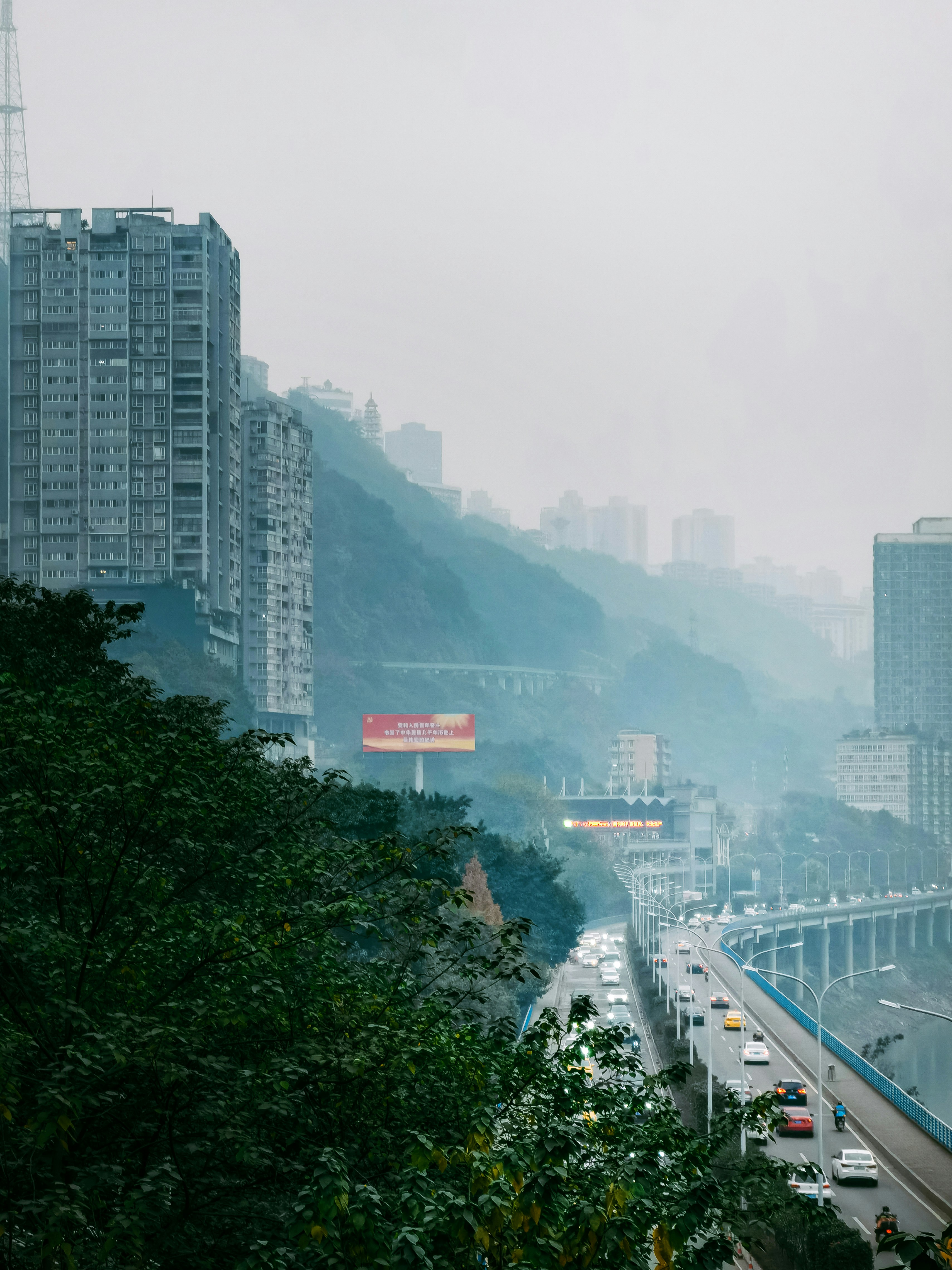 Dense cityscape with a freeway curling along the hillside and tall buildings receding into blue-gray haze. A bright red billboard punctuates the scene, drawing the eye amid the mist.