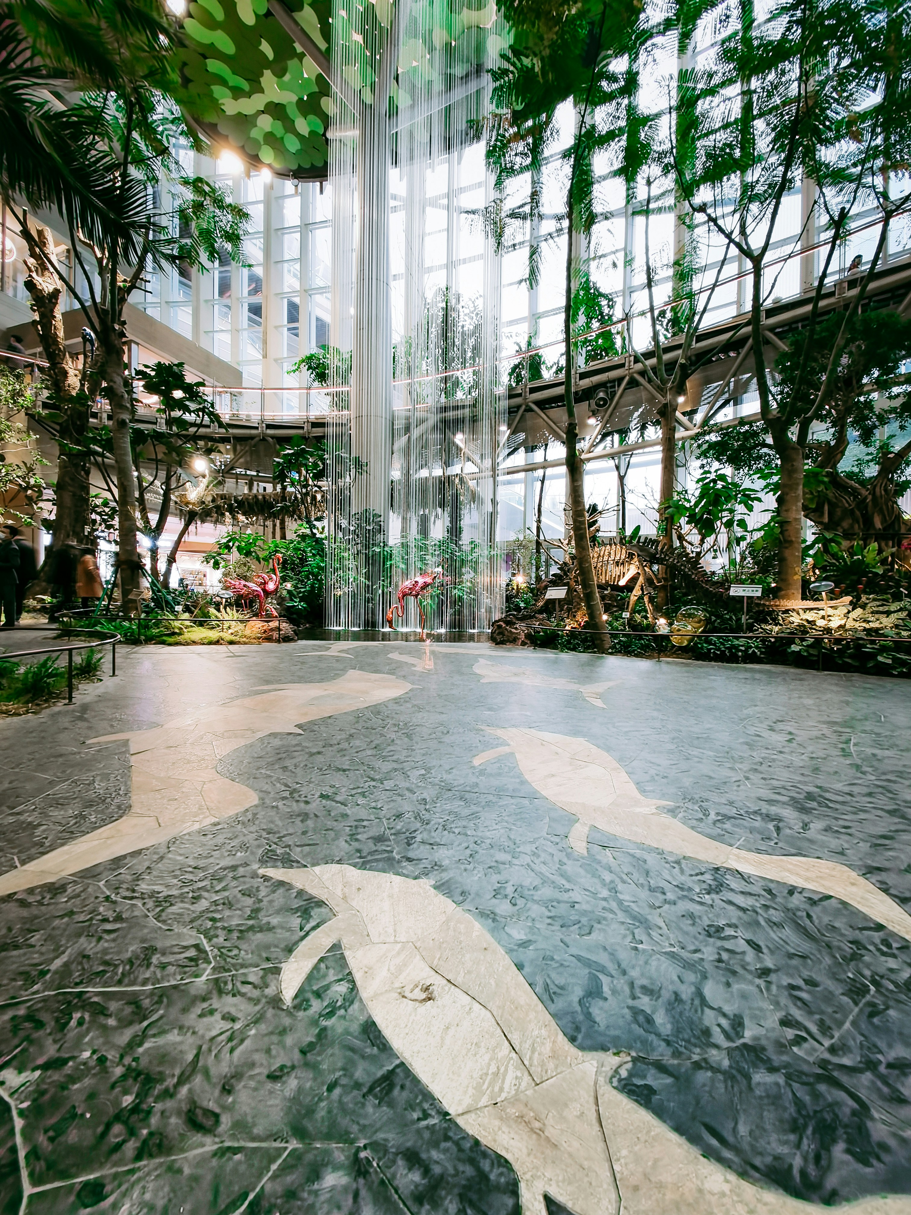 Lush indoor rainforest atrium with a tall glass waterfall feature and patterned marble floor.
