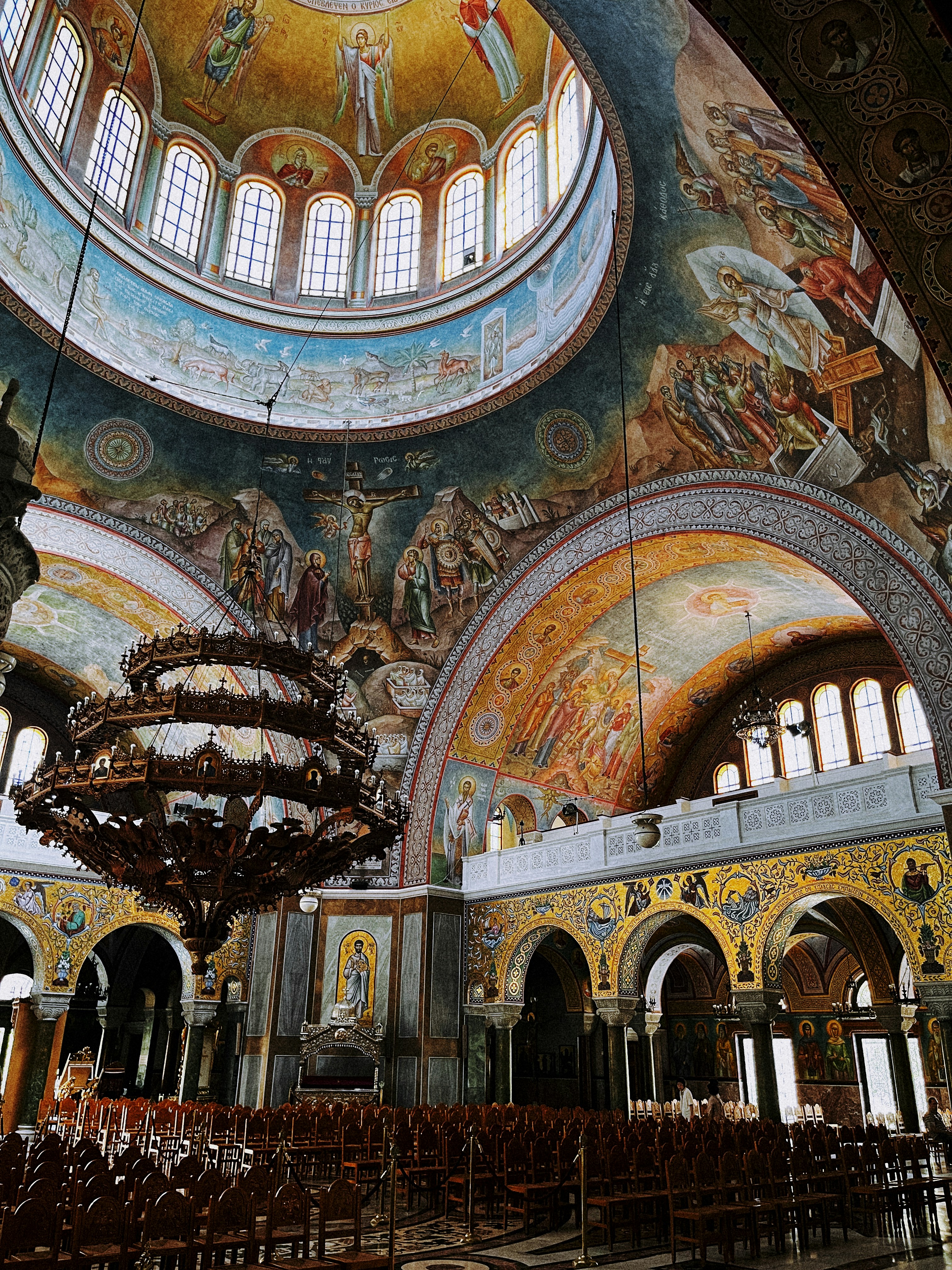 Ornate Byzantine cathedral interior featuring a grand wooden chandelier and frescoed domes. Rows of wooden pews line the nave beneath arched galleries.