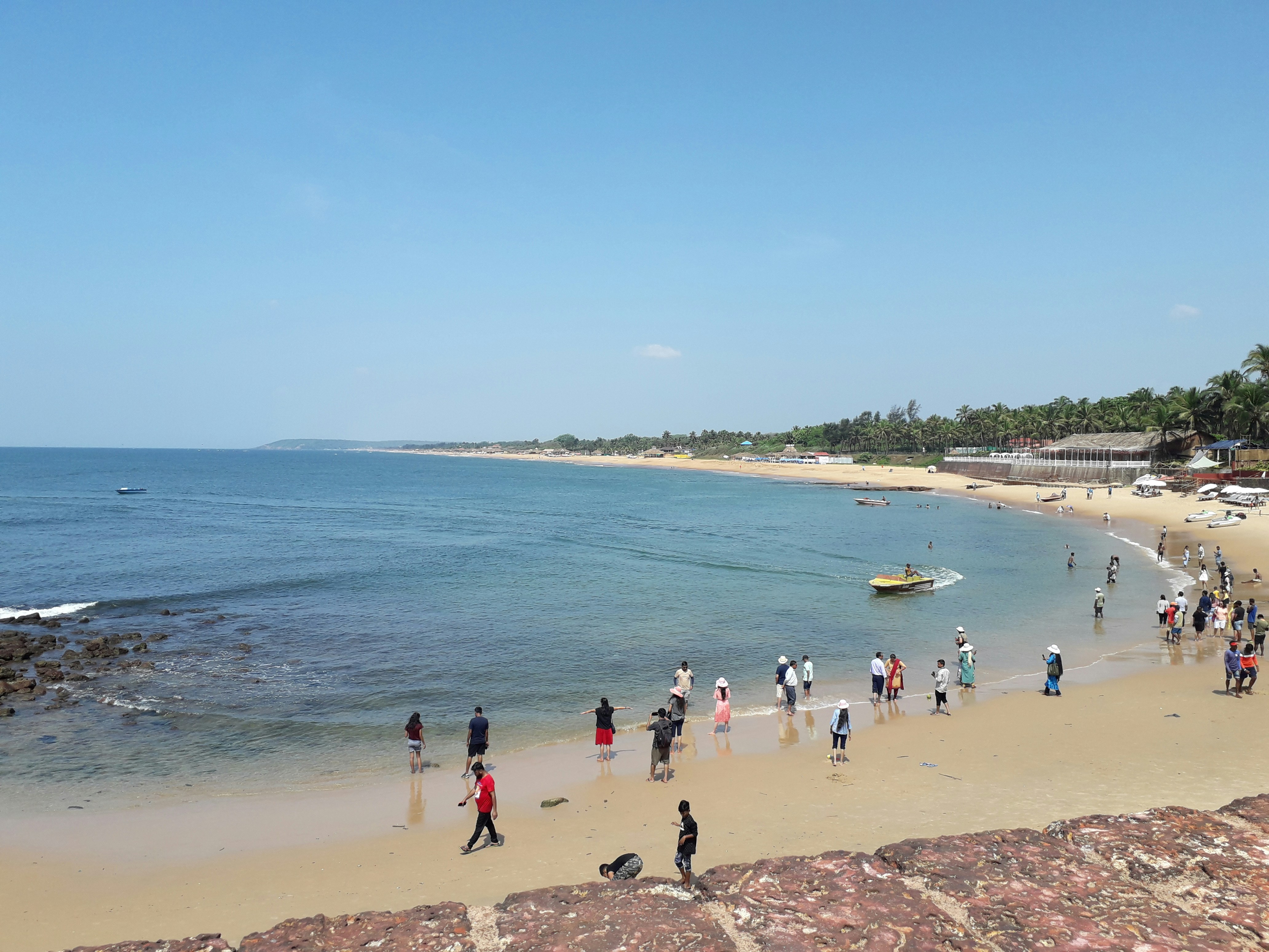 Sunlit crescent beach crowded with families and beachgoers along the shore. A small boat rests offshore against a calm blue sea. Palm trees line the distant horizon.