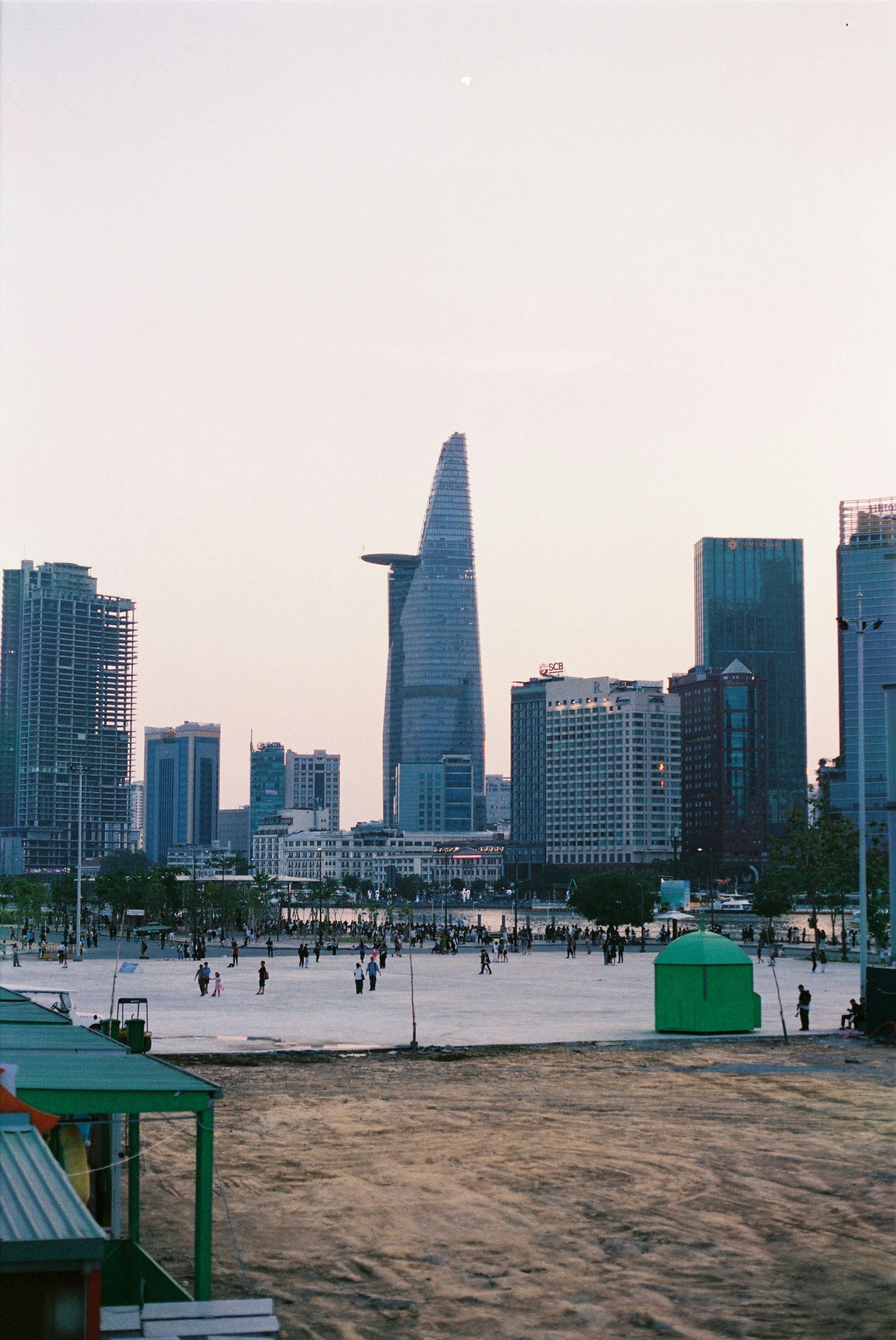 Urban waterfront skyline with the Bitexco Financial Tower among high-rise buildings, seen from a sandy foreground with people and a green shelter.