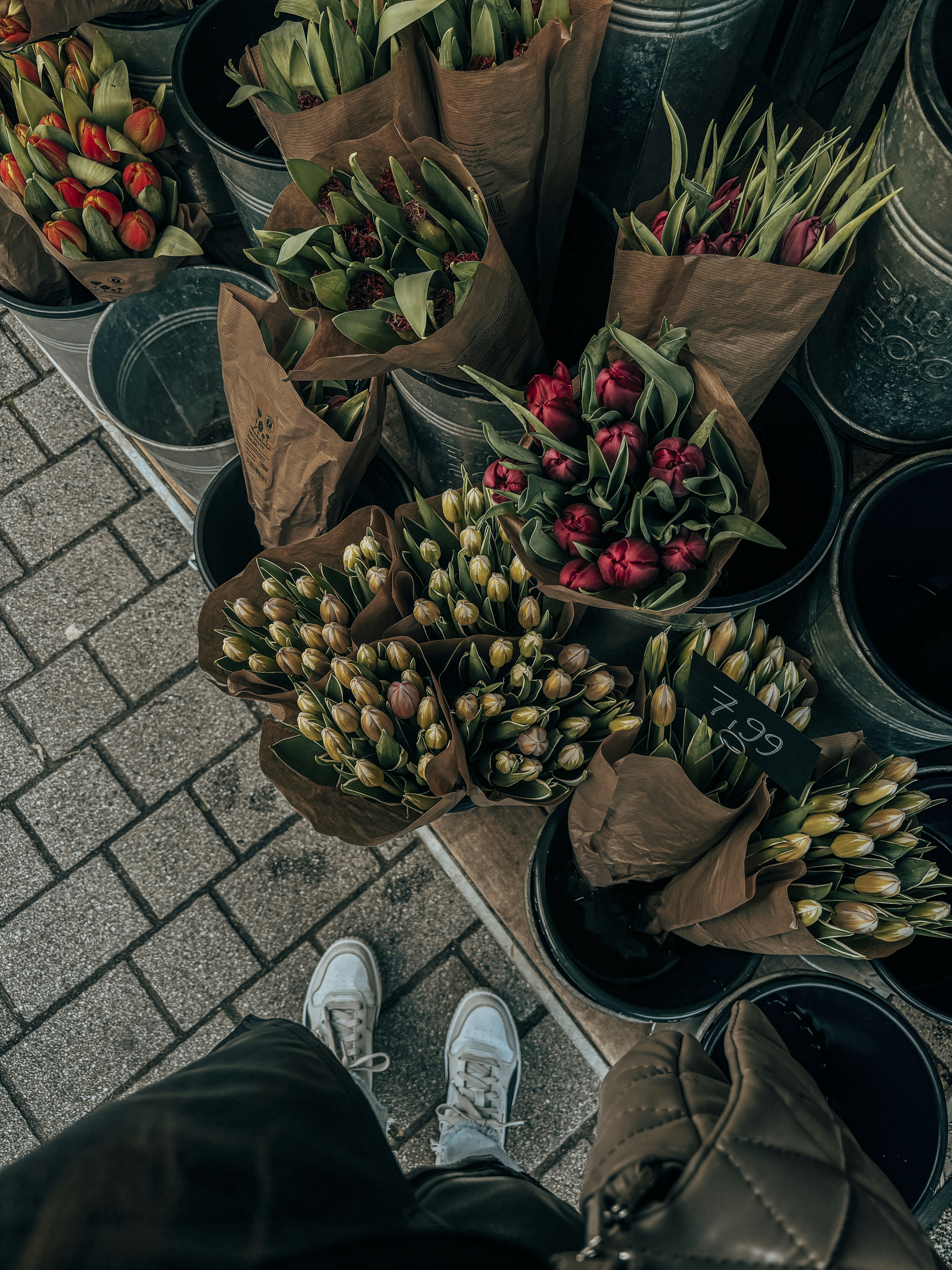 Top-down photograph of a flower market: bouquets of tulips wrapped in brown paper arranged in metal buckets on a cobblestone sidewalk, with sneakers at the bottom edge.
