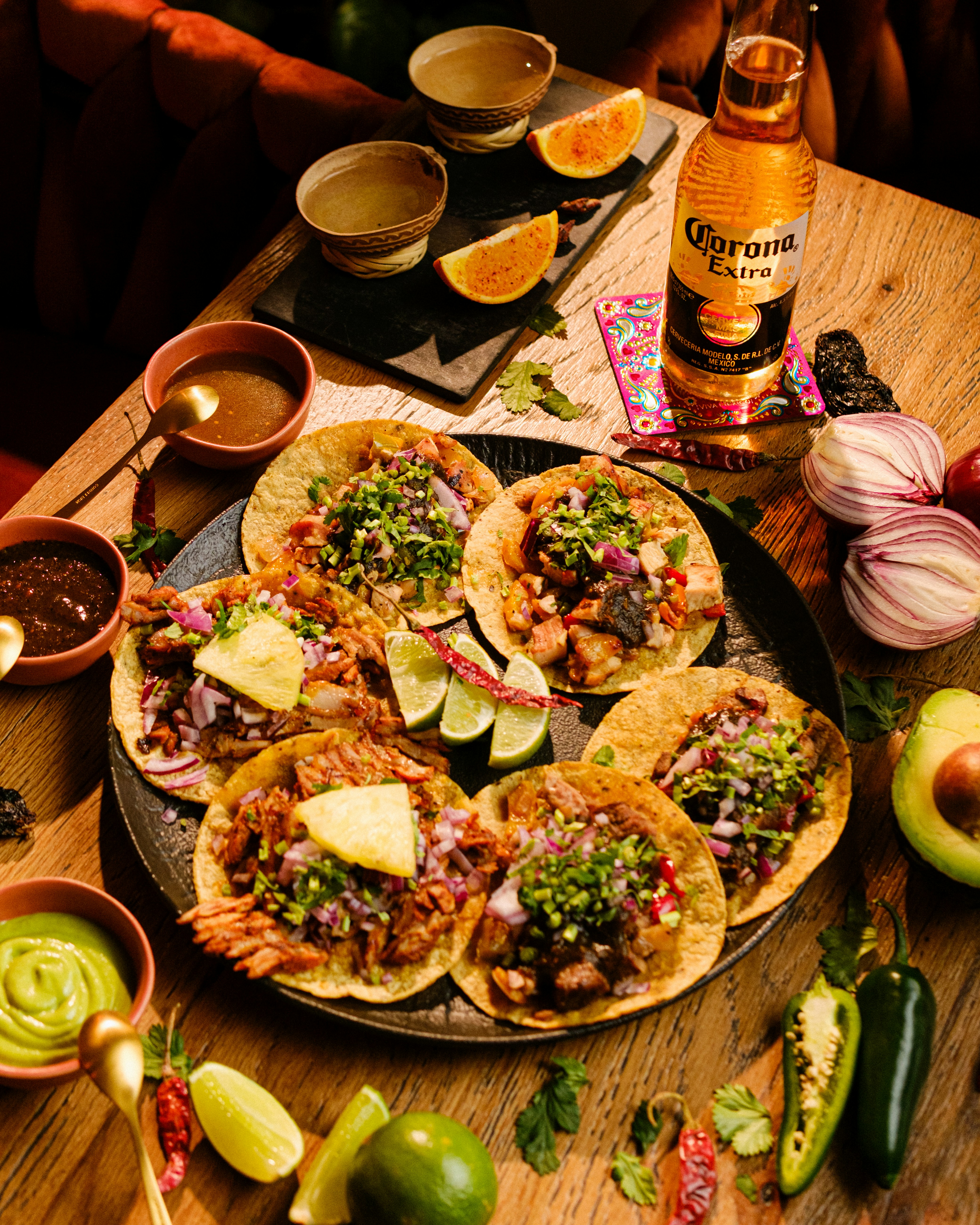 Vibrant taco platter with lime wedges, chopped onions, and cilantro rests on a wooden table beside bowls of salsa and a Corona bottle. The composition conveys a warm, festive dining scene.