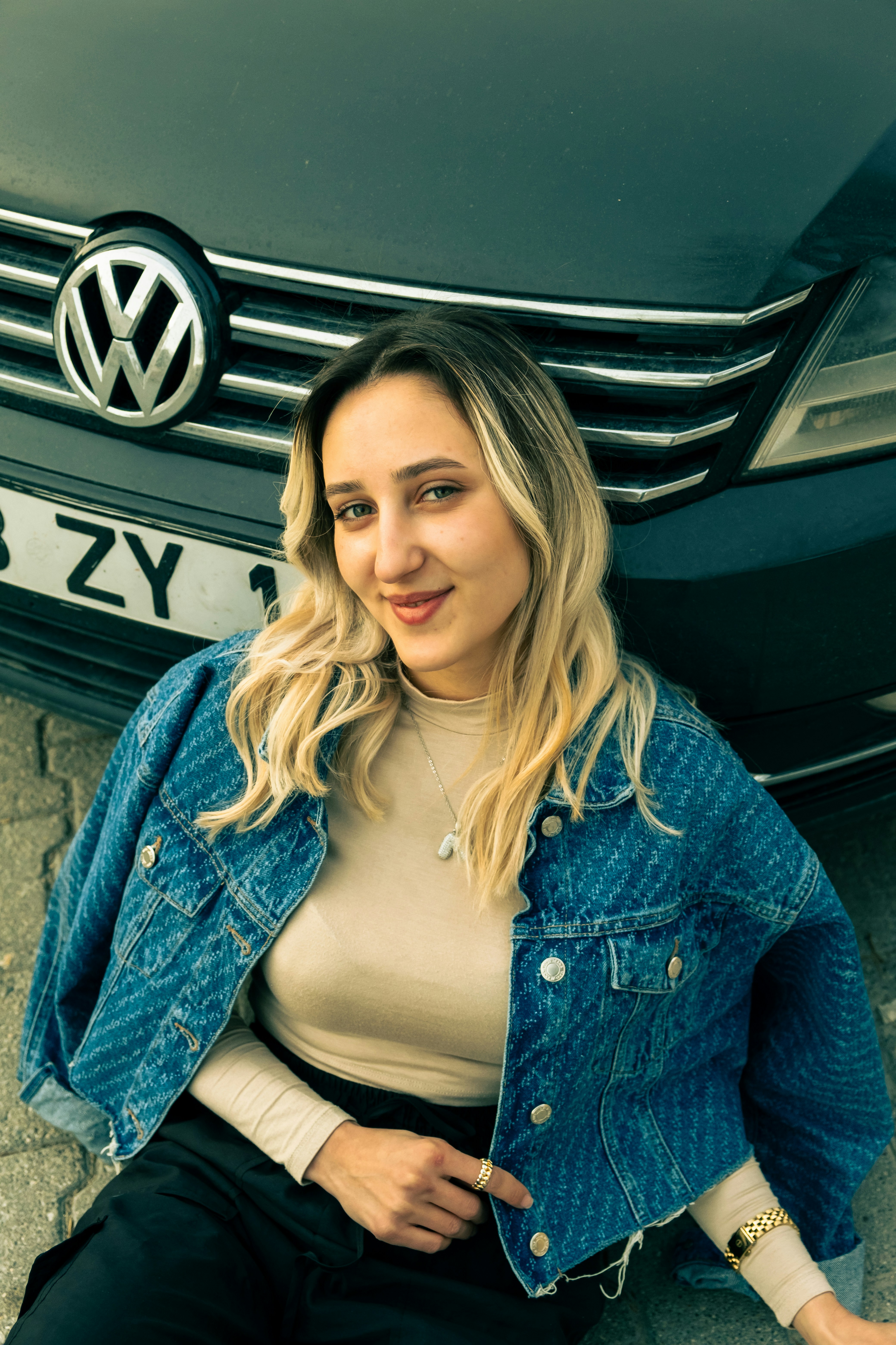 A young woman poses casually in front of a Volkswagen car, showcasing her stylish denim jacket and relaxed demeanor.