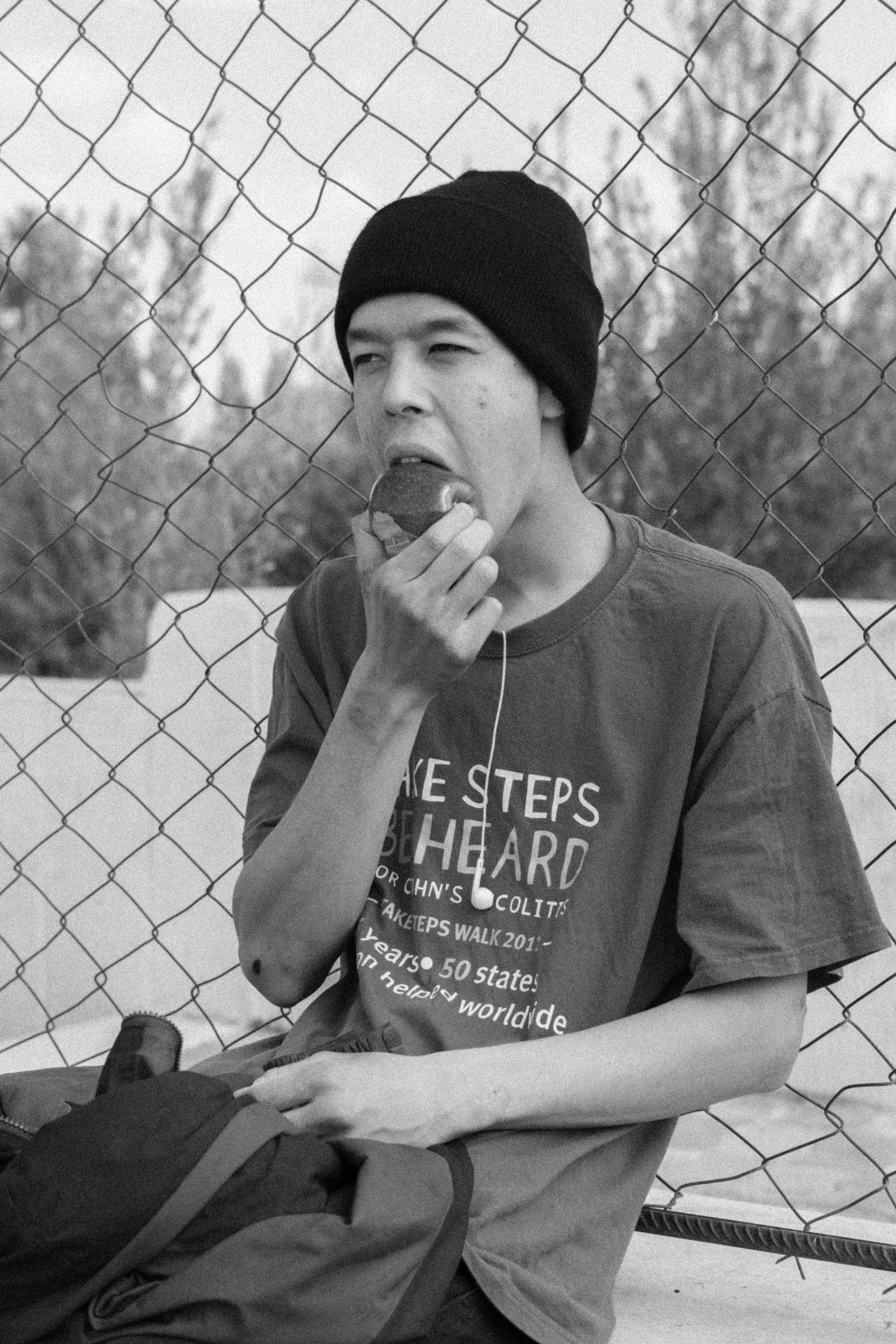 Young man in a beanie enjoying a snack, seated near a chain-link fence, with trees softly blurred in the background.