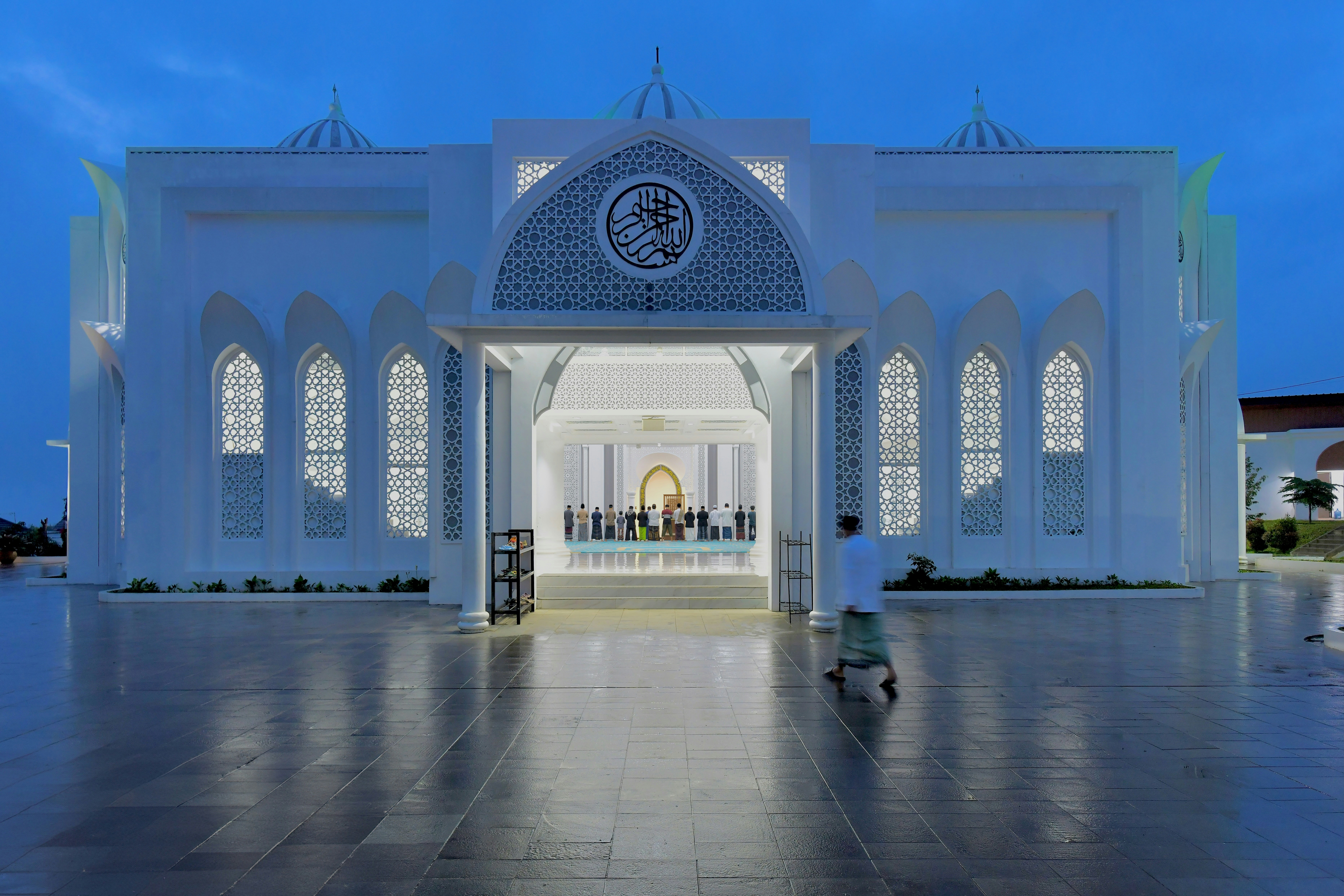 Illuminated mosque facade reflecting on wet pavement during blue hour.