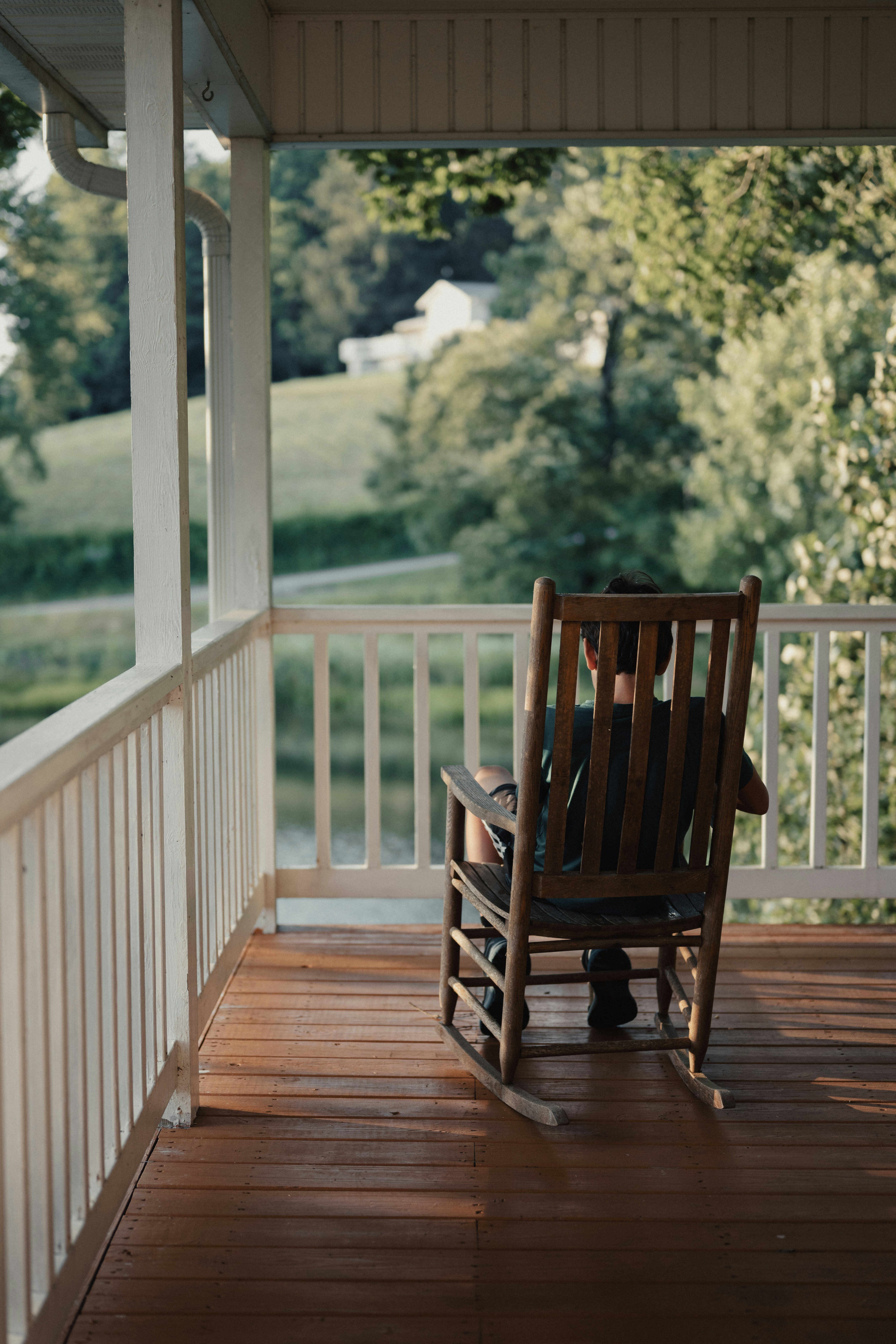 A person sits in a rocking chair on a porch, gazing out at a serene landscape filled with trees and distant buildings.