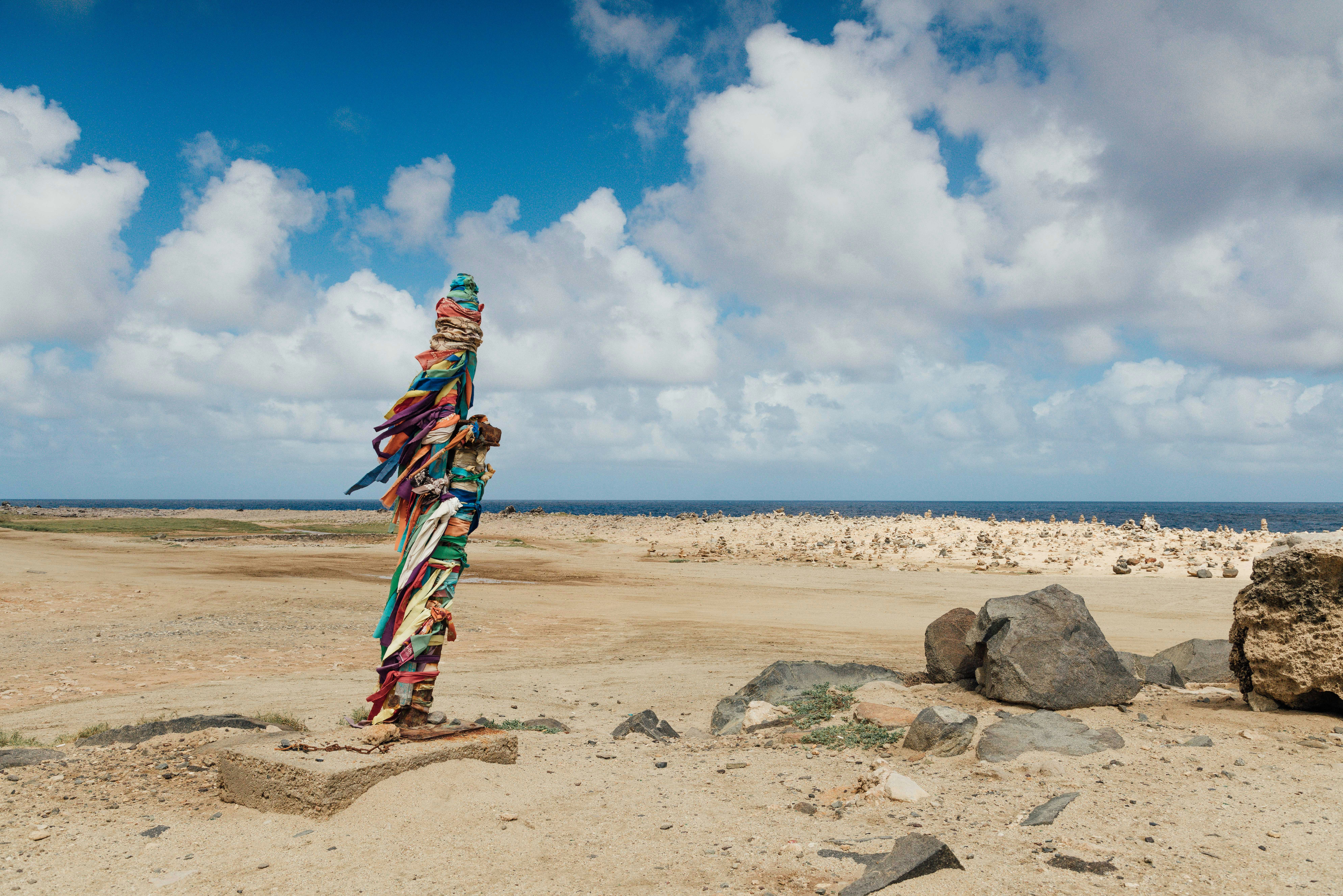 Colorful ribbons tied to a wooden post stand against a vast coastal landscape, with rocky formations and a serene ocean backdrop. The scene captures the essence of cultural expression in a natural setting.