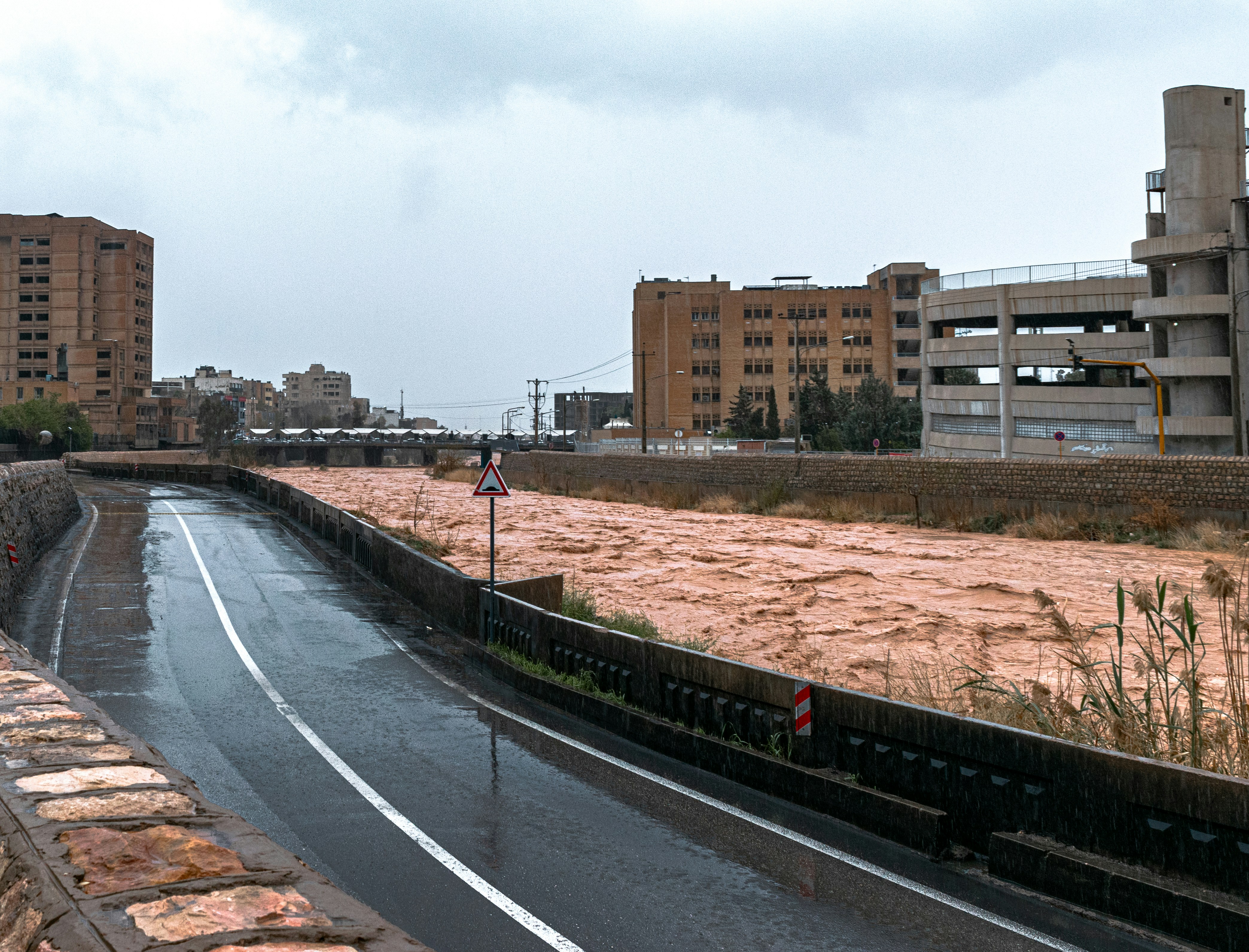 Flooded river rushing alongside city buildings under a cloudy sky.