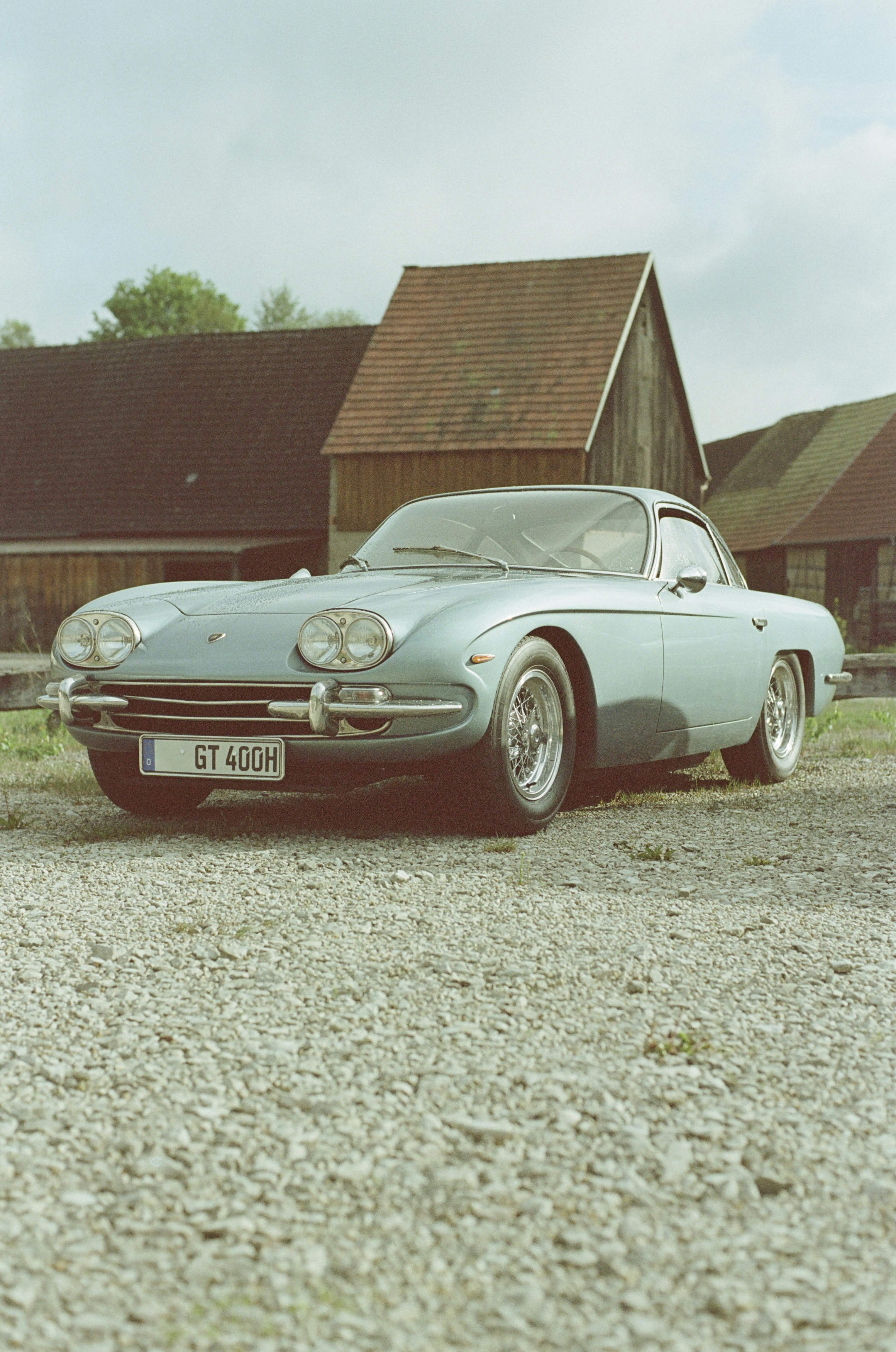 Light blue Lamborghini 400 GT parked on gravel with rustic barns in the background.