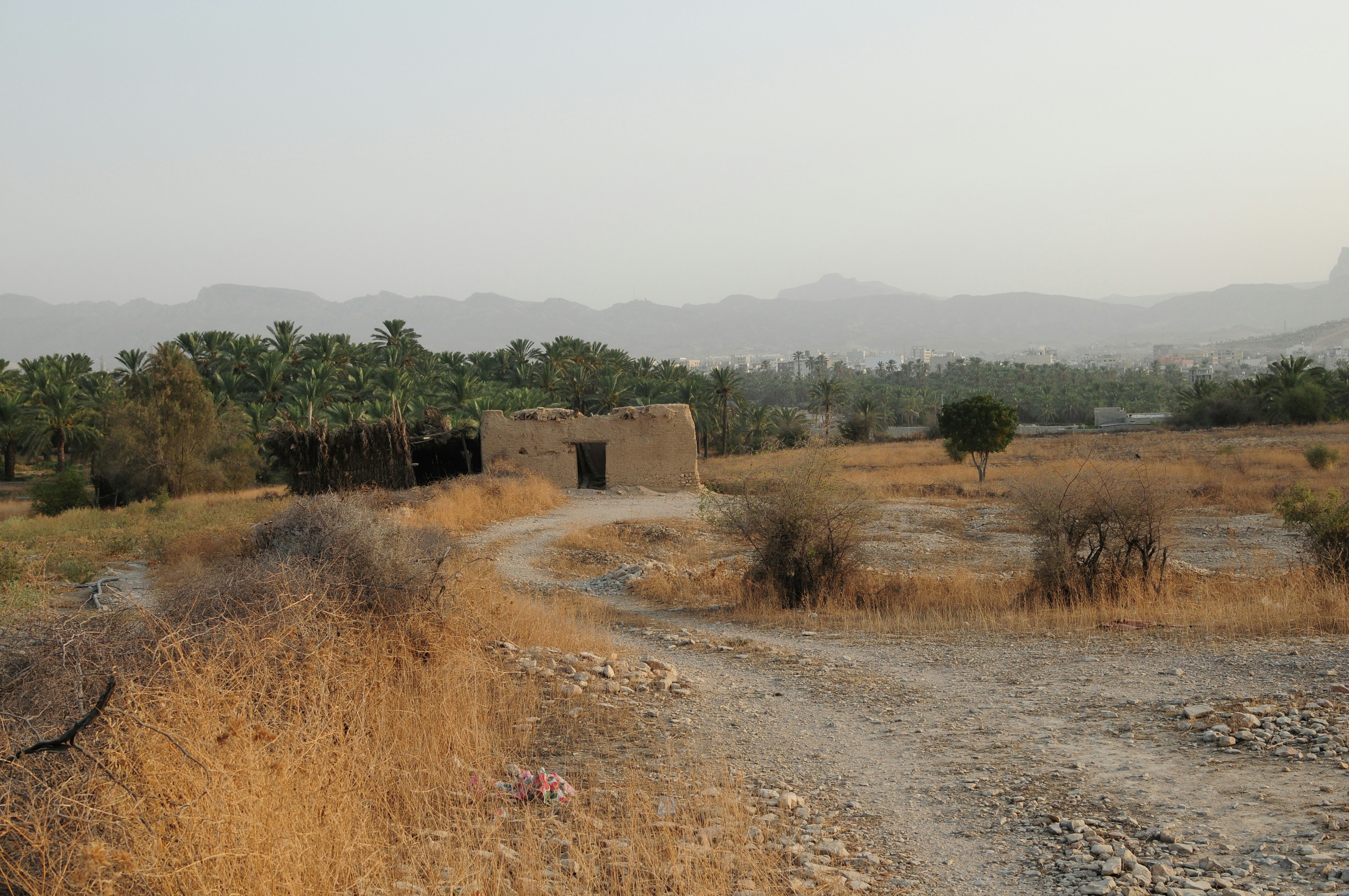 Dirt path leading to a rustic stone building in a dry landscape with distant mountains.