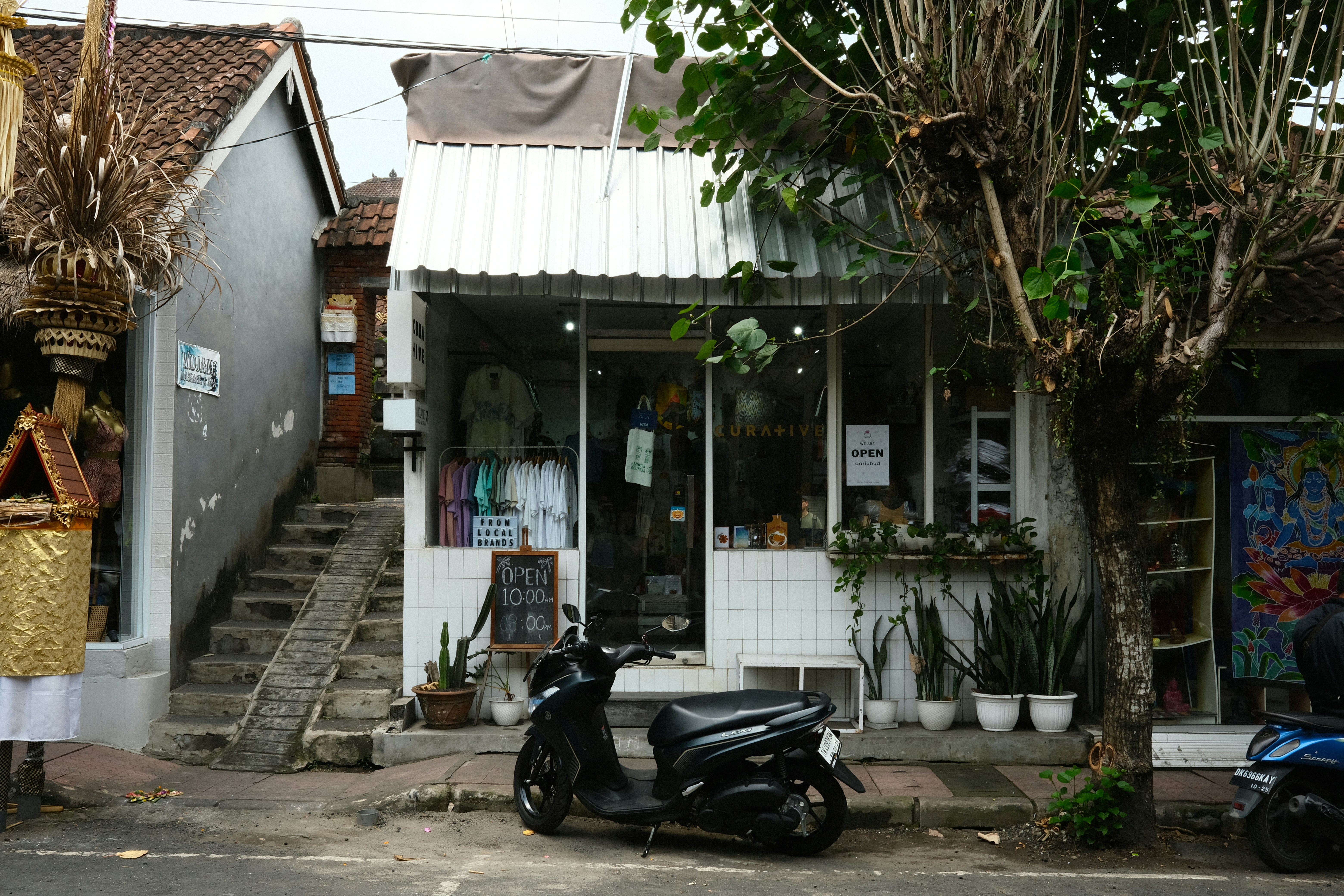 Motor scooter parked outside a quaint boutique with potted plants and a colorful display under a tree-lined street.