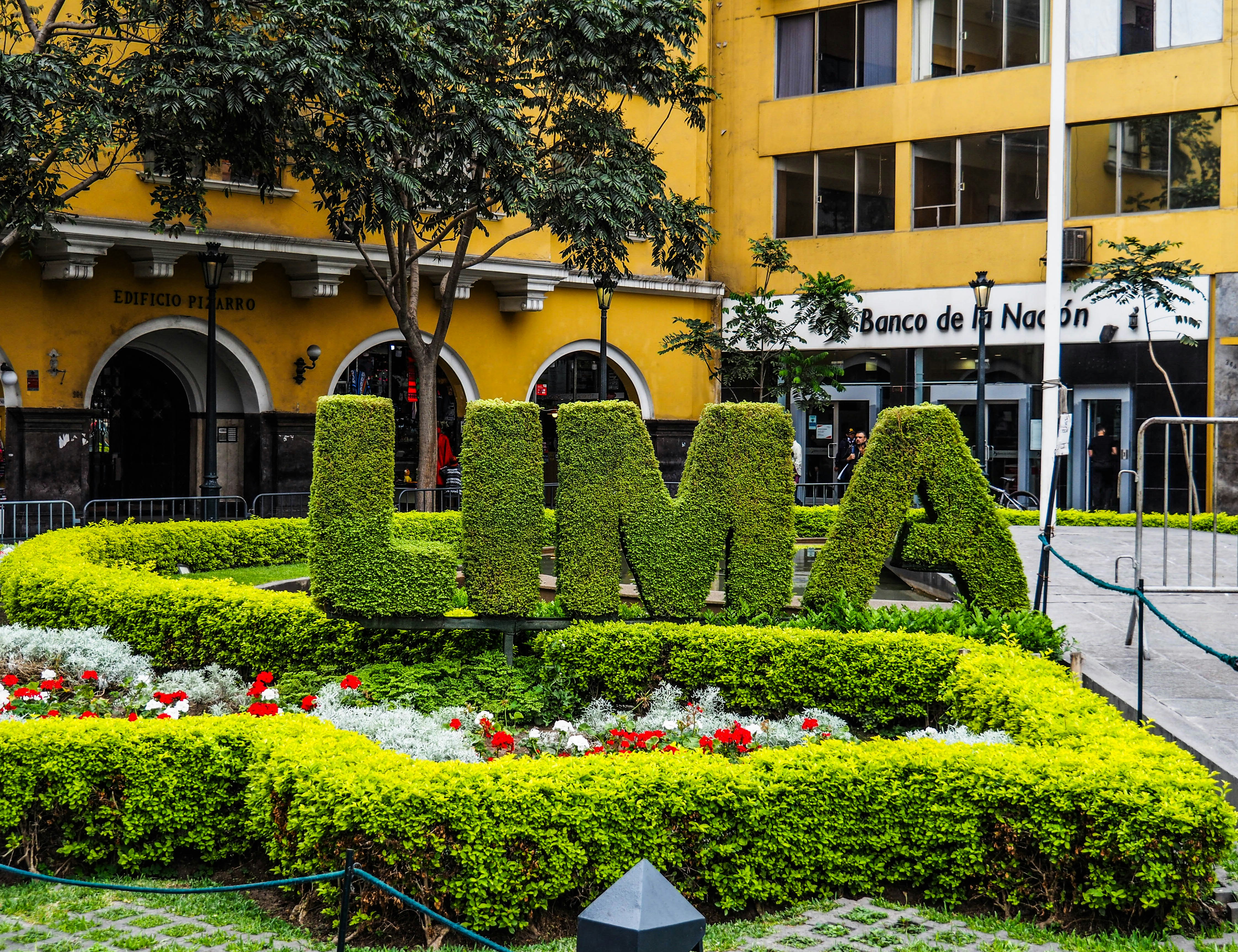 Topiary letters spelling 'LIMA' amidst vibrant flower beds in a public square, showcasing the city's charm.