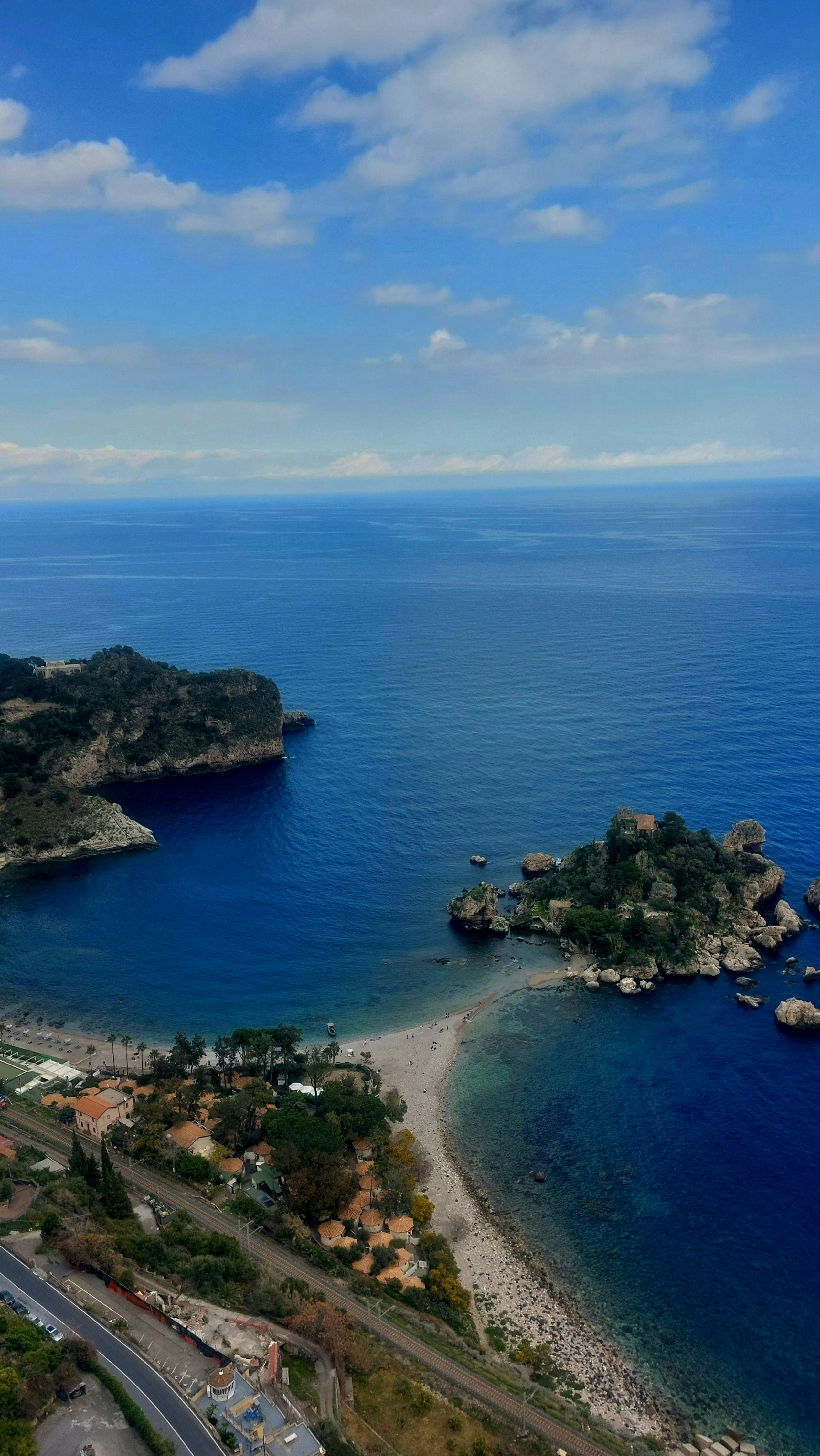 Vast blue sea meets a rocky coastline with a small village along the shore, captured from an aerial perspective showing water, rocks, and coastal road layout.