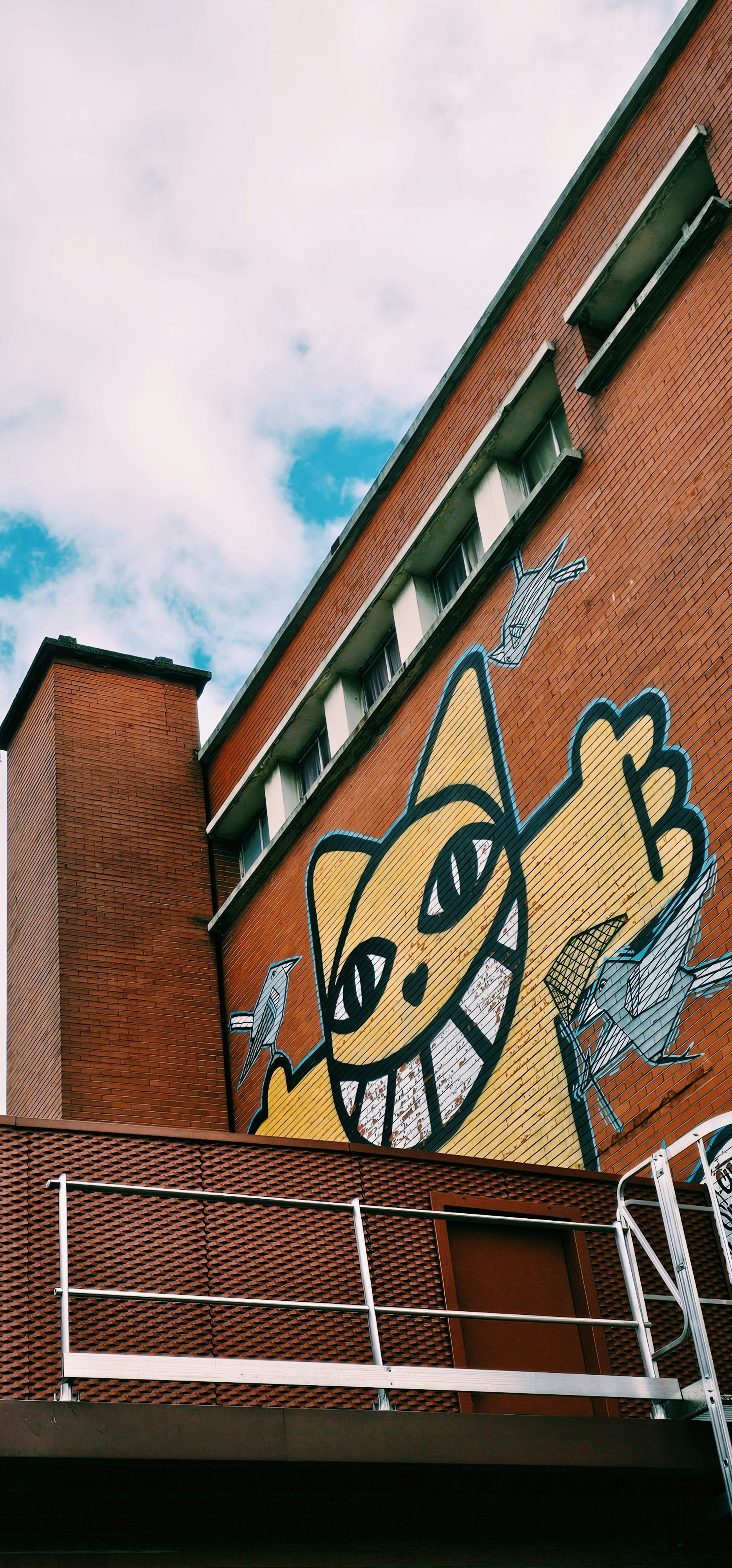 Vibrant graffiti painting of a smiling yellow cat on a red brick wall, with a metal railing in the foreground and a blue sky overhead.