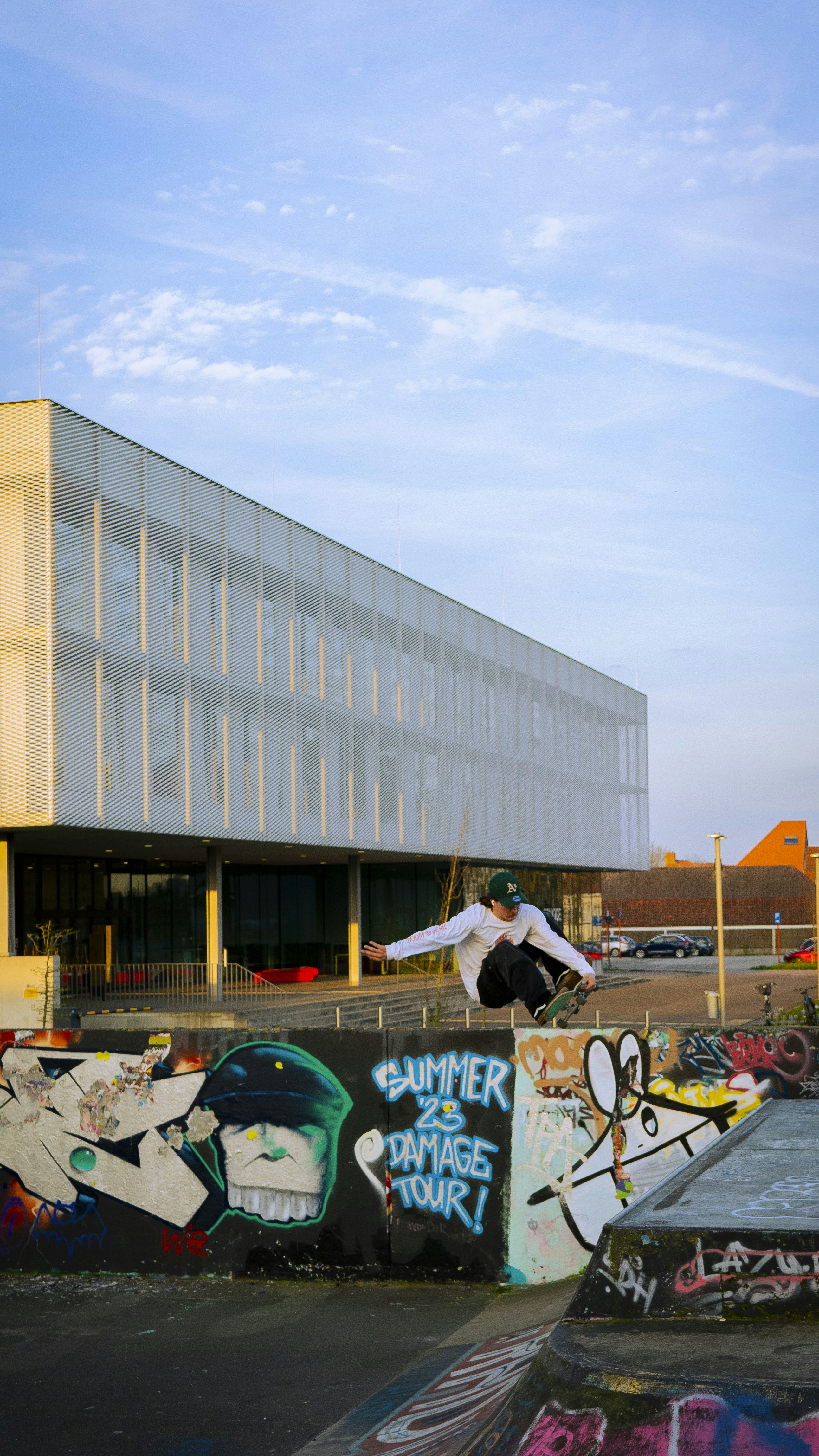 Skateboarder performing a trick over a graffiti-covered ramp in an urban skate park, with a modern building in the background.