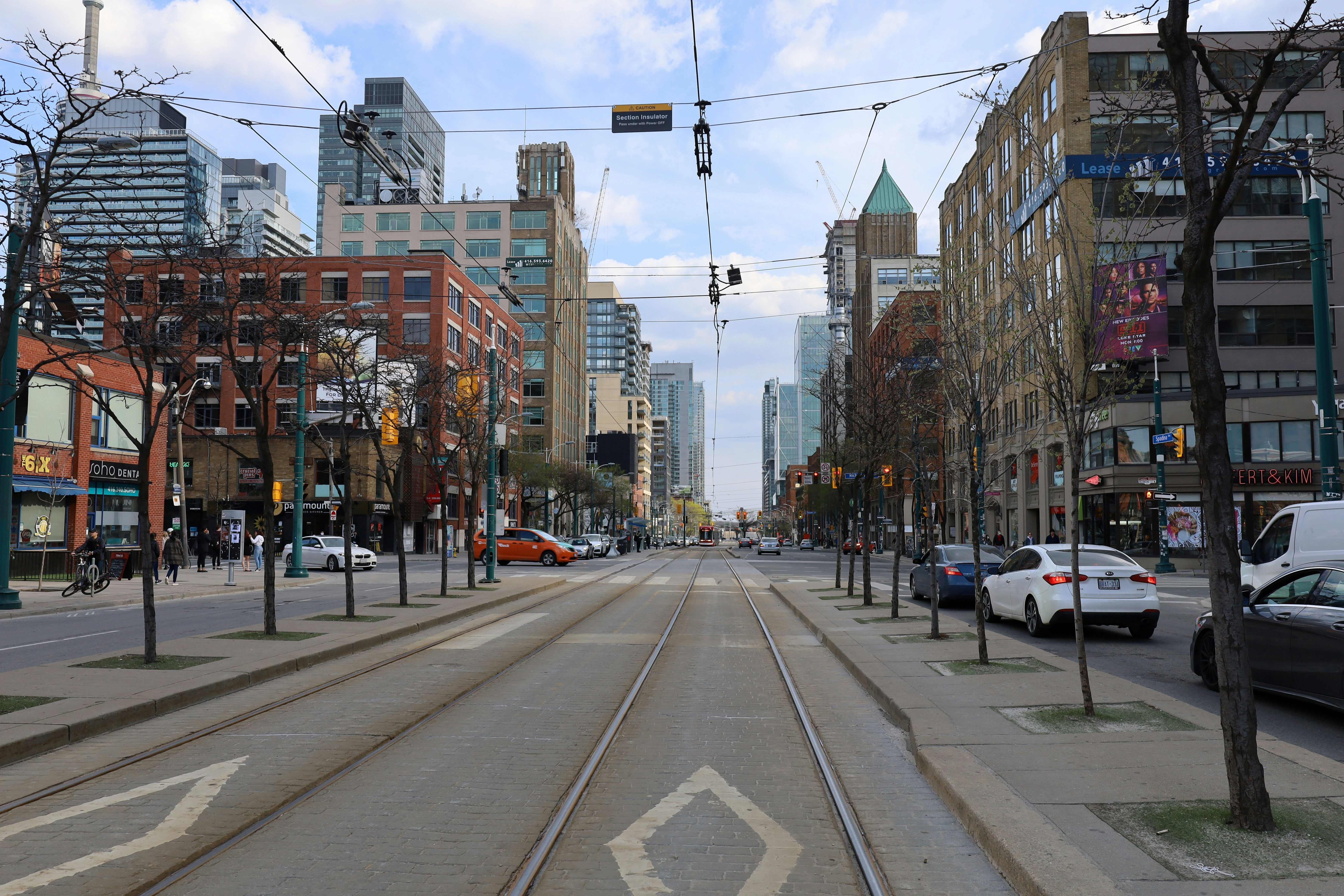 Tram tracks leading through a bustling urban landscape, showcasing a blend of modern architecture and city life. Trees line the street, emphasizing the contrast between nature and city structures.