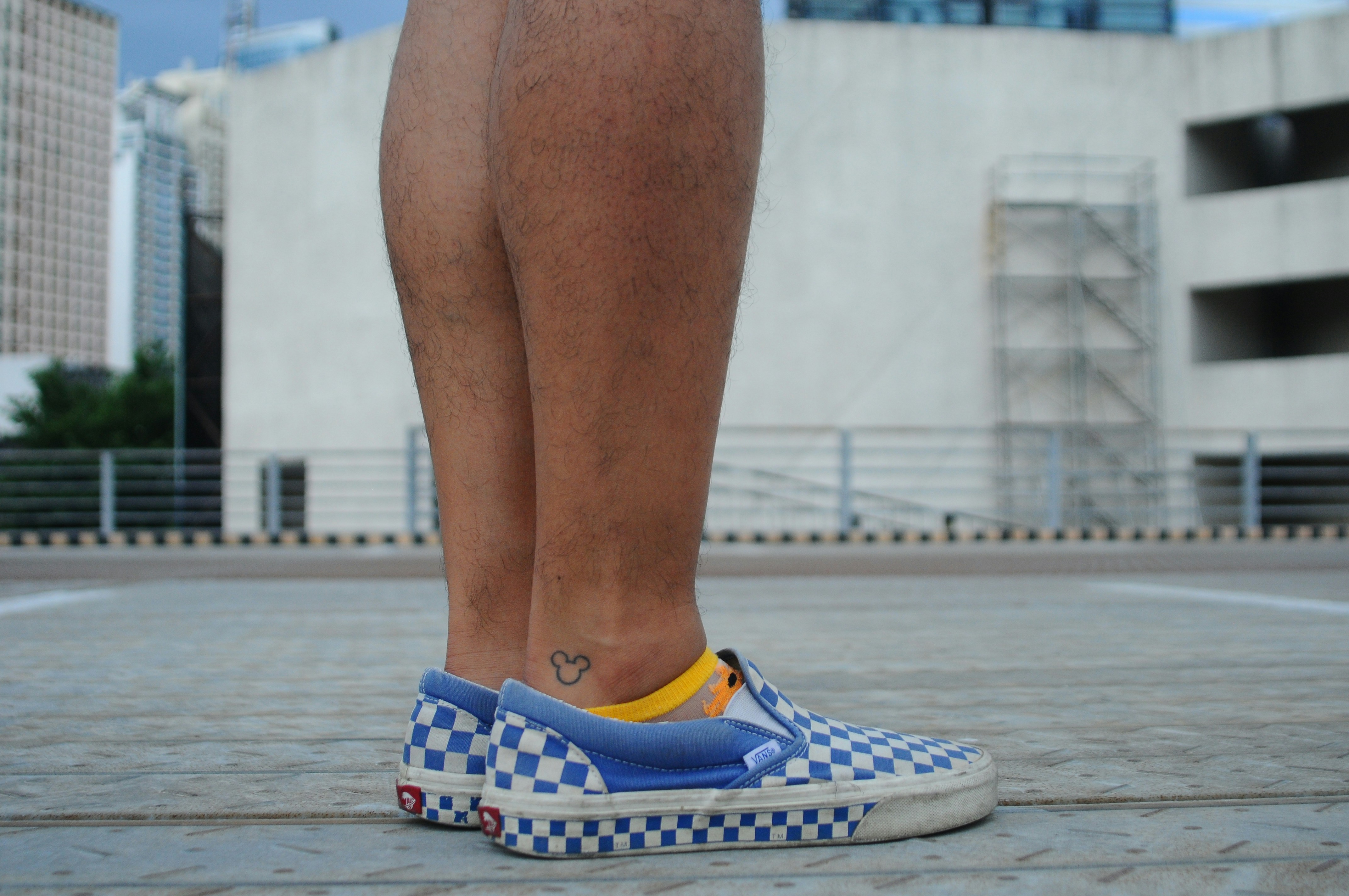 Person wearing checkered sneakers with a visible ankle tattoo, standing on a concrete surface.