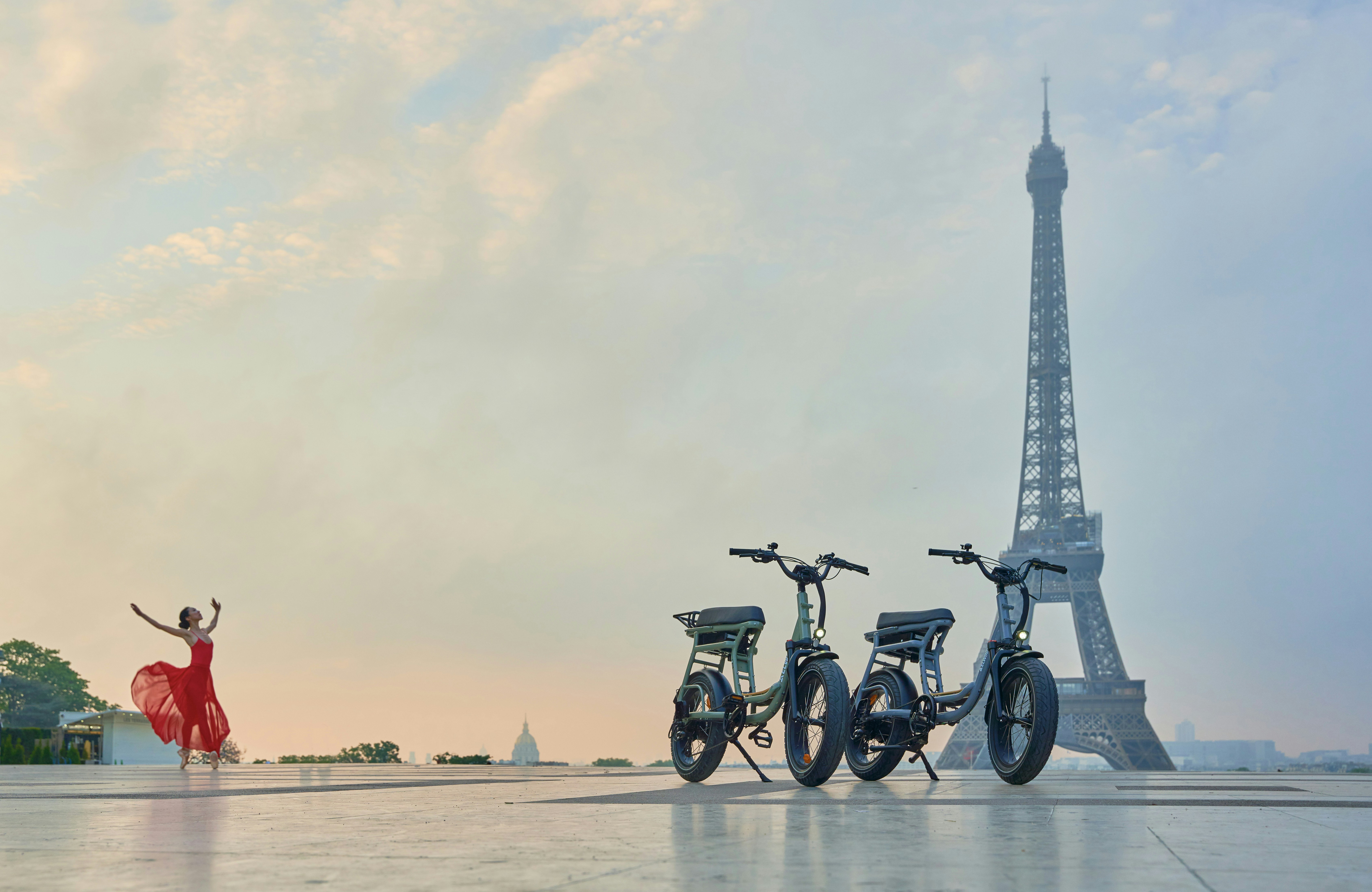 A woman wearing a red dress dancing on a square facing a beautiful view on the Eiffel tower in Paris, at sunrise, next to two electric bikes beautifully parked.