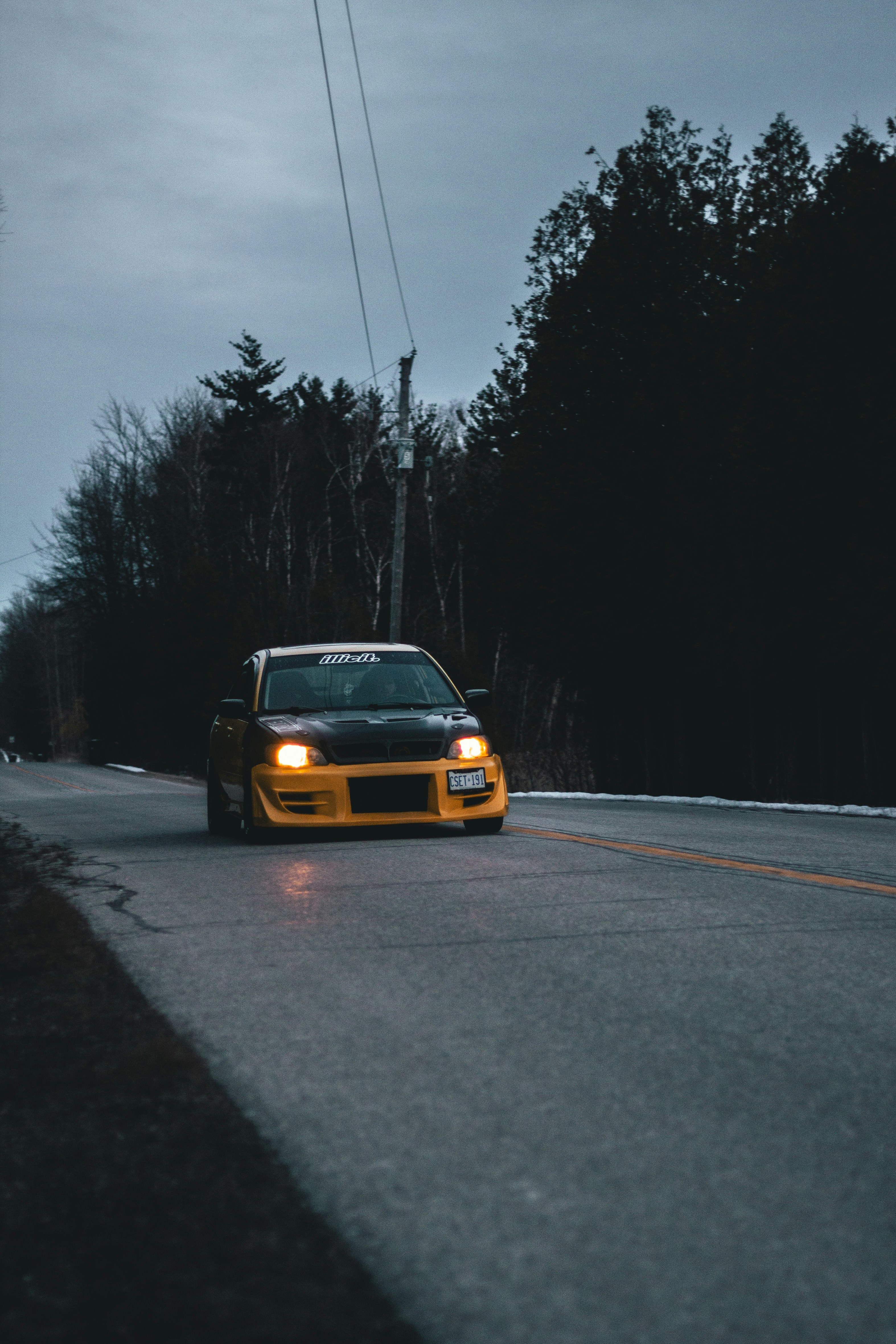 A yellow car driving down a road next to a forest photo – Free Car ...