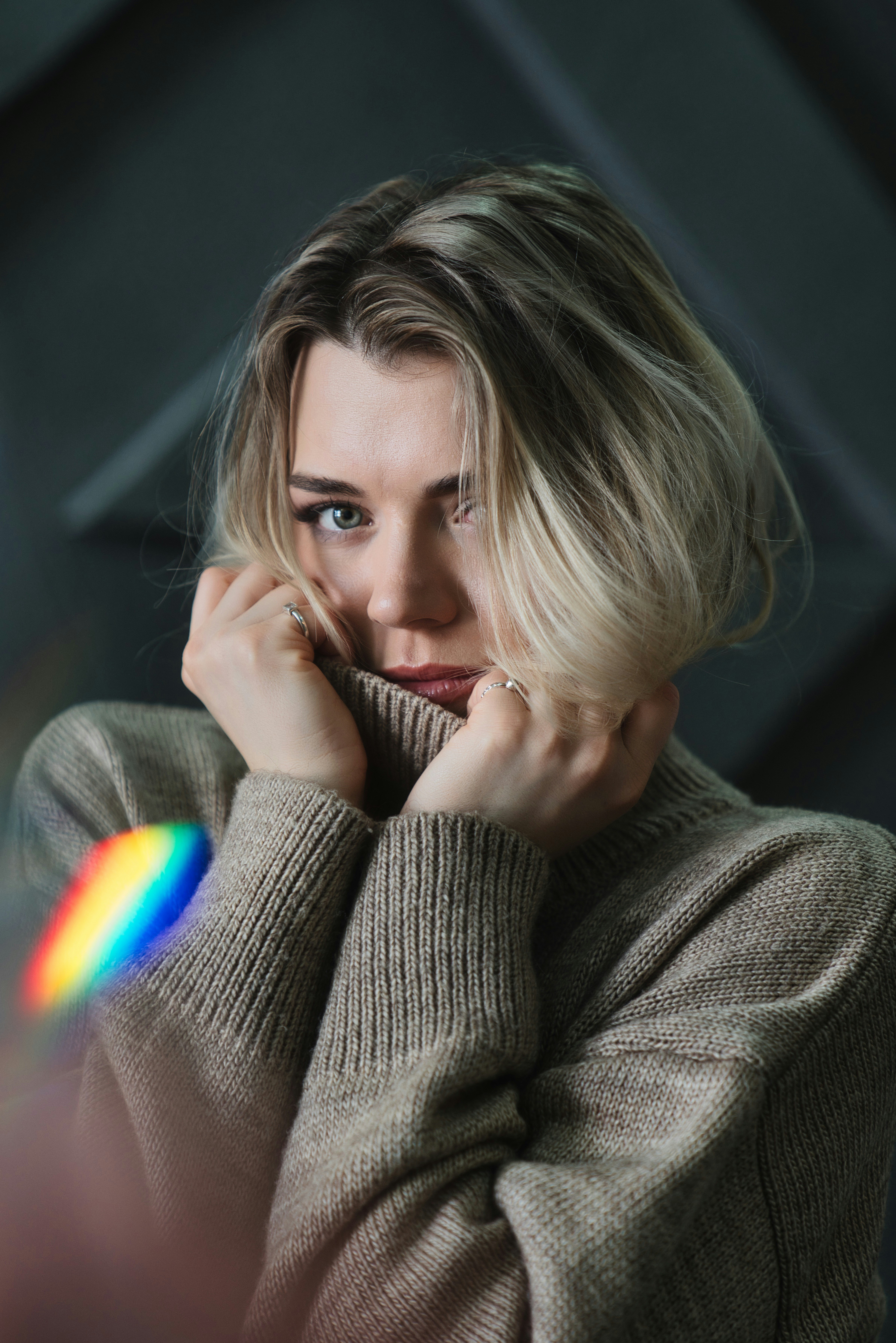 Portrait of a woman in a beige knit sweater, meeting the camera with a soft, intimate gaze. A rainbow prism blur in the foreground adds colorful contrast.