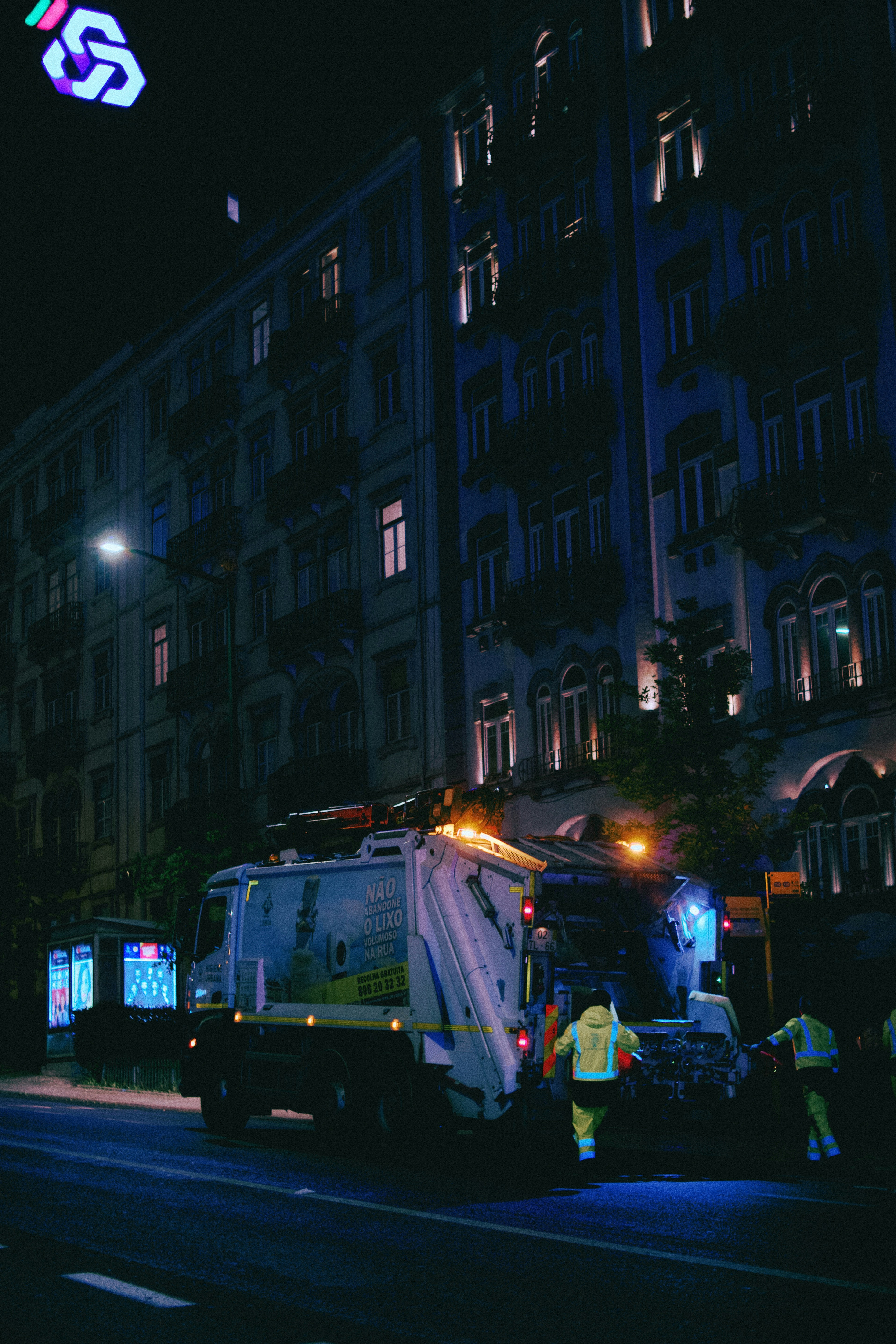 Garbage truck and sanitation workers engaged in nighttime waste collection along a city street, illuminated by streetlights and building facades.