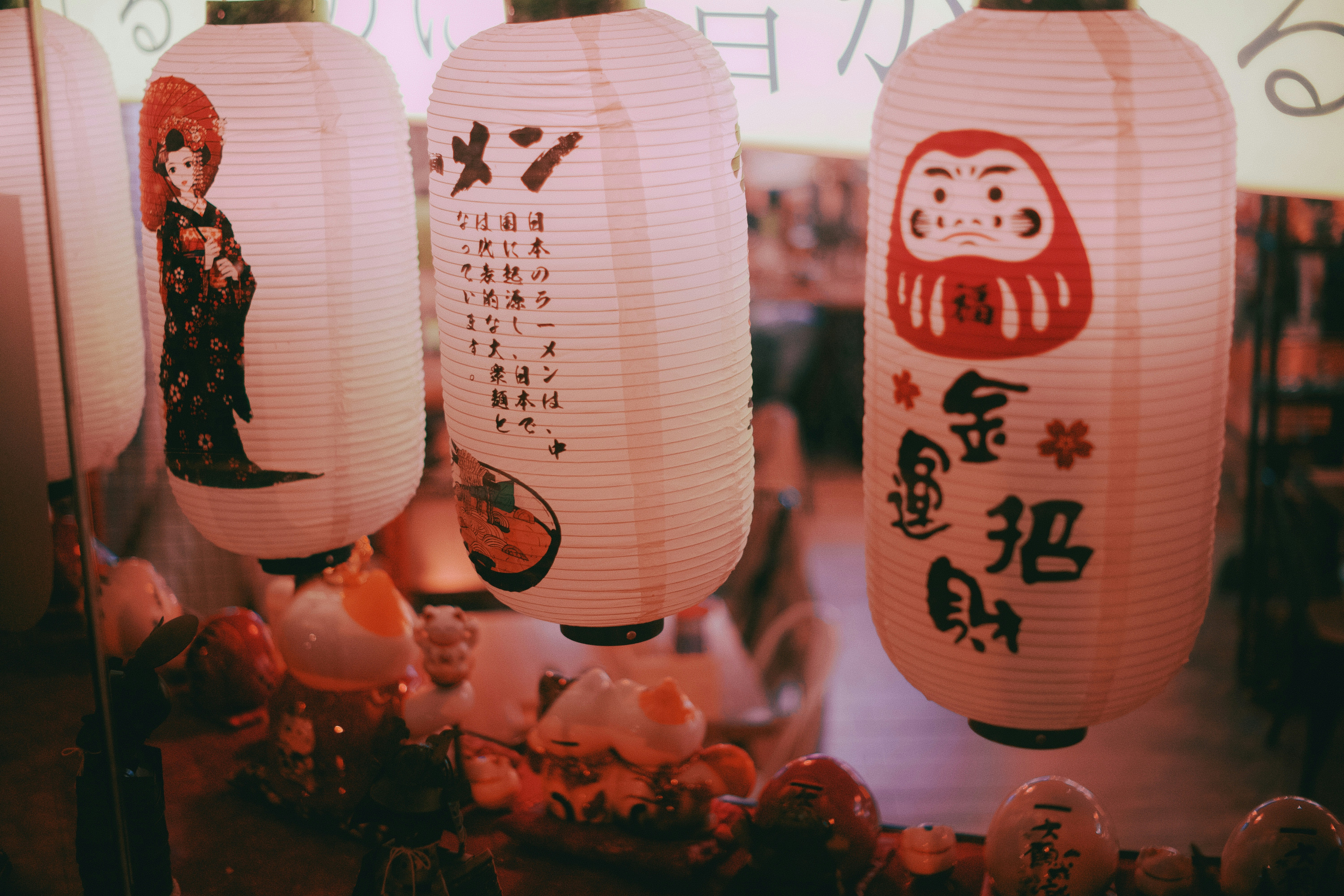 Warm paper lanterns glow inside a glass storefront, with miniature figurines and kanji inscriptions along the fronts.