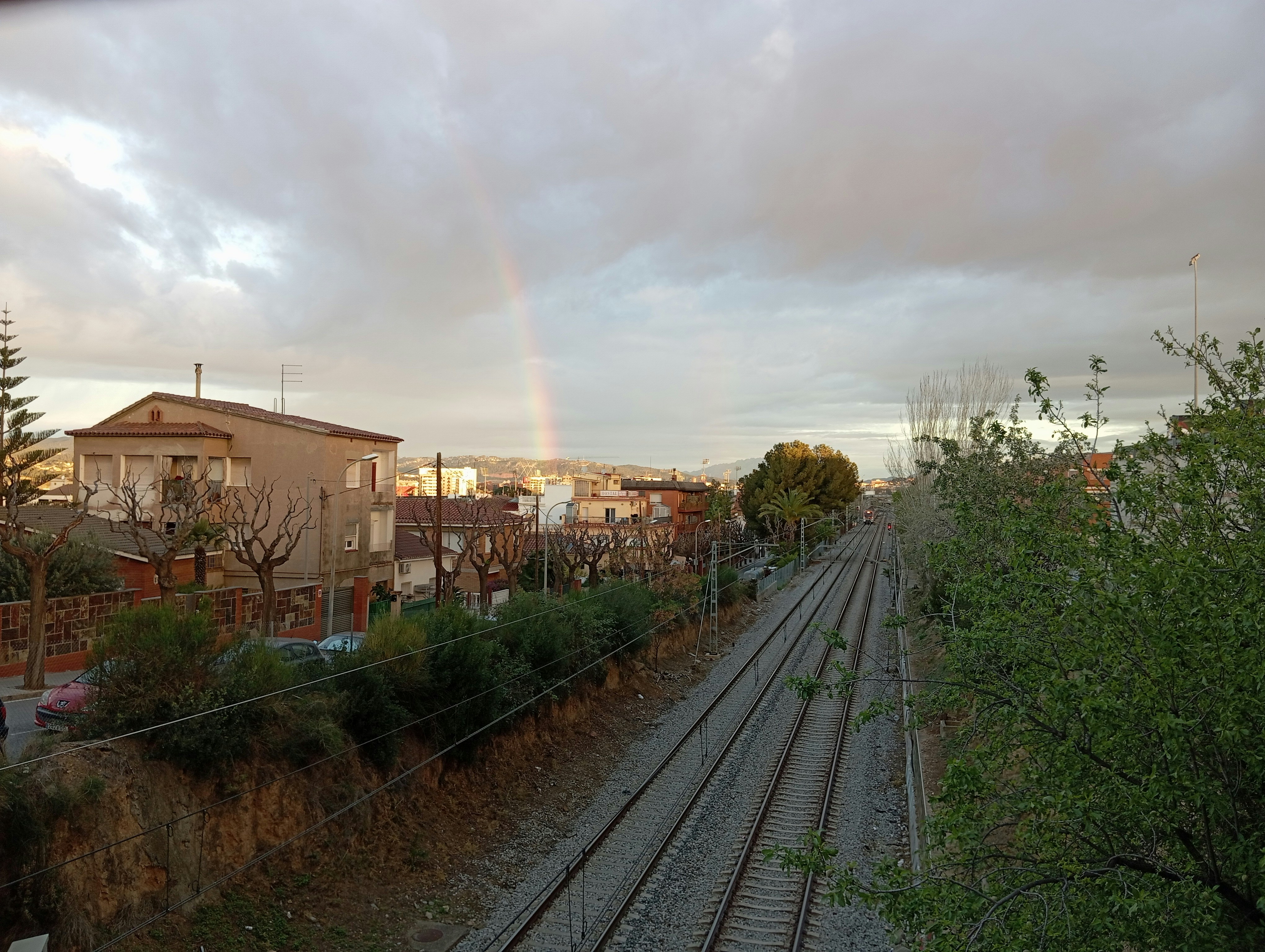 Rainbow arches over suburban houses and railway tracks at dusk.