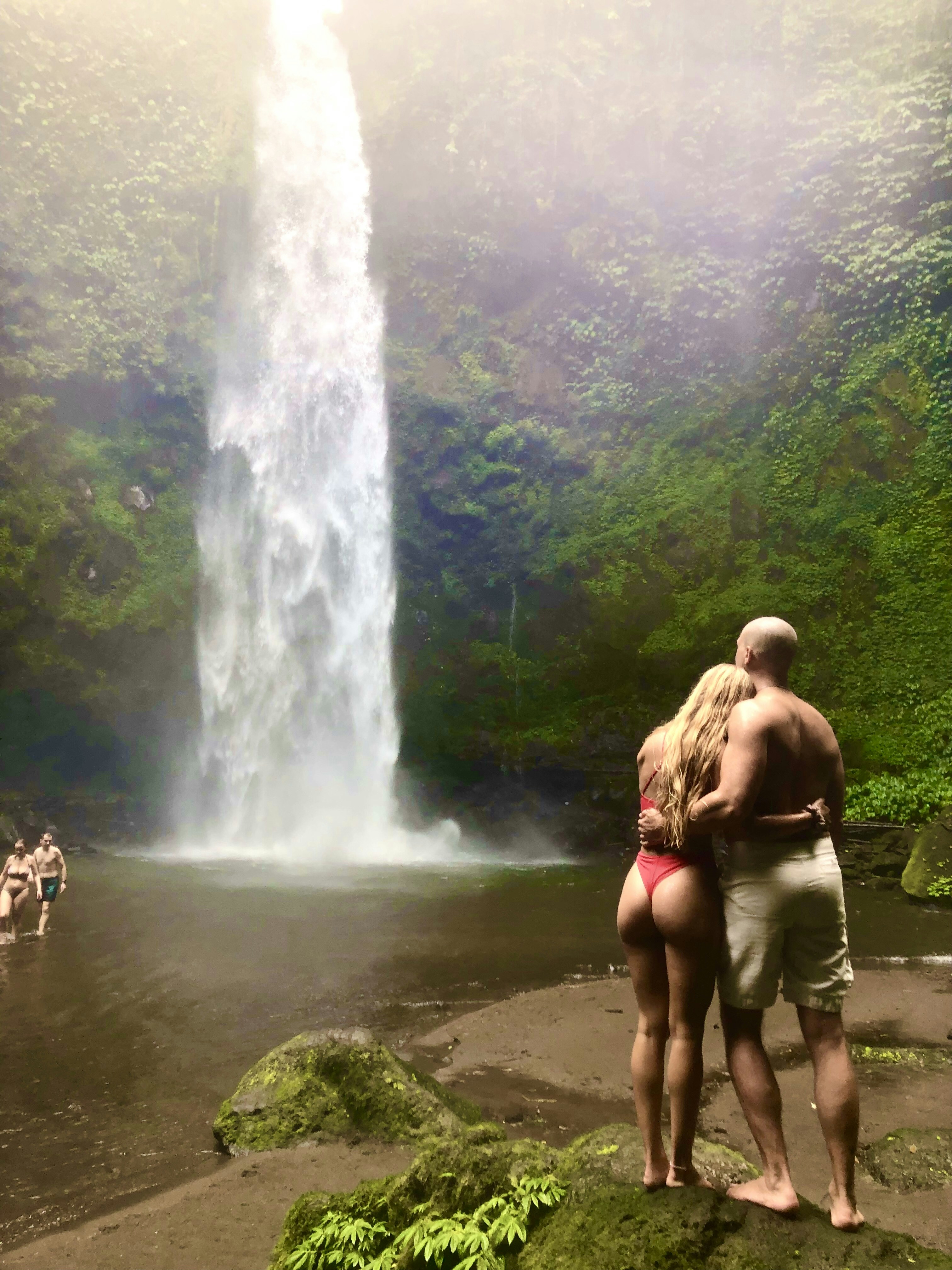 Couple embracing in front of a majestic waterfall surrounded by lush greenery. The scene captures the intimacy of their moment against the backdrop of cascading water.