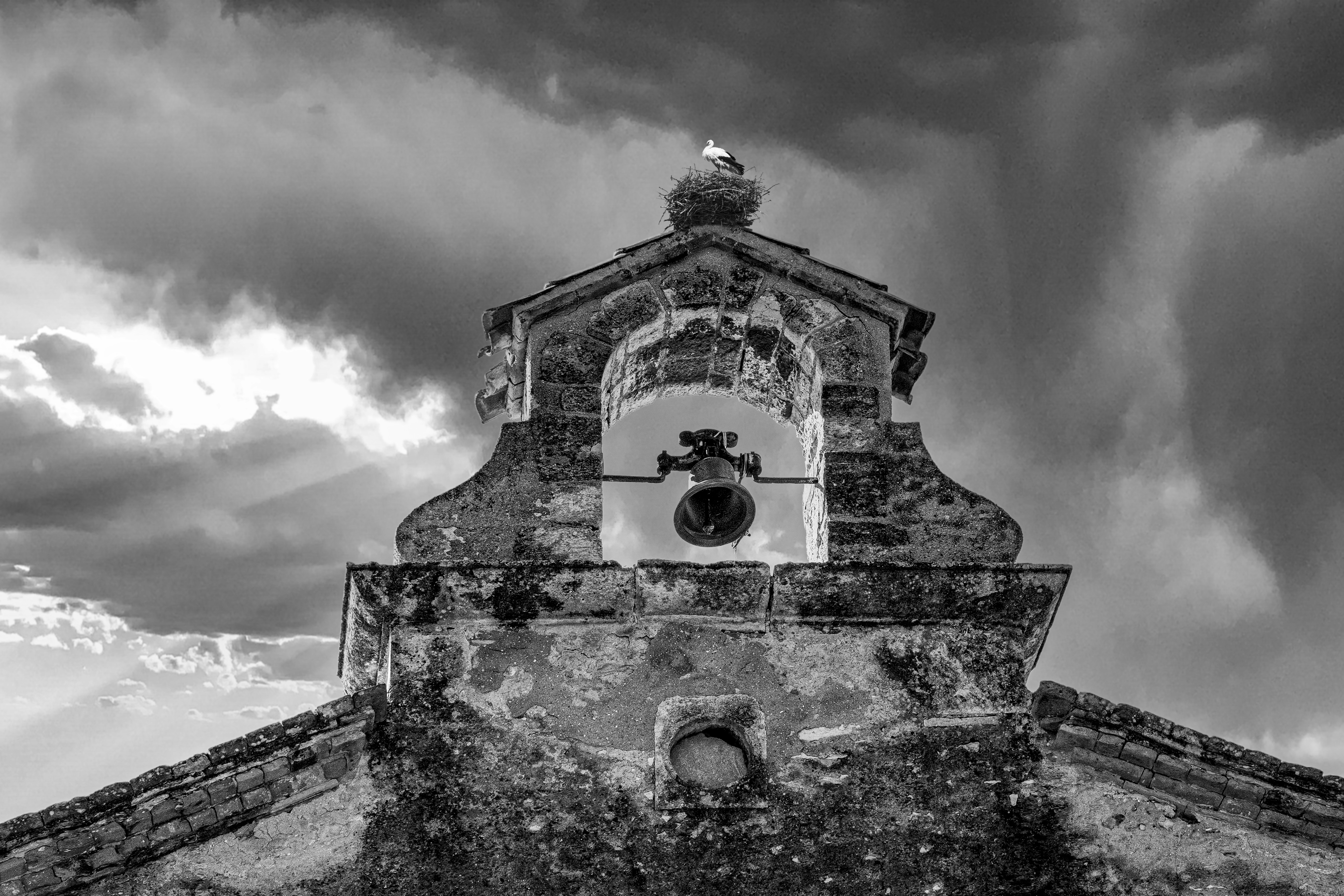 Black and white photo of a weathered stone bell tower under dramatic, cloudy skies.