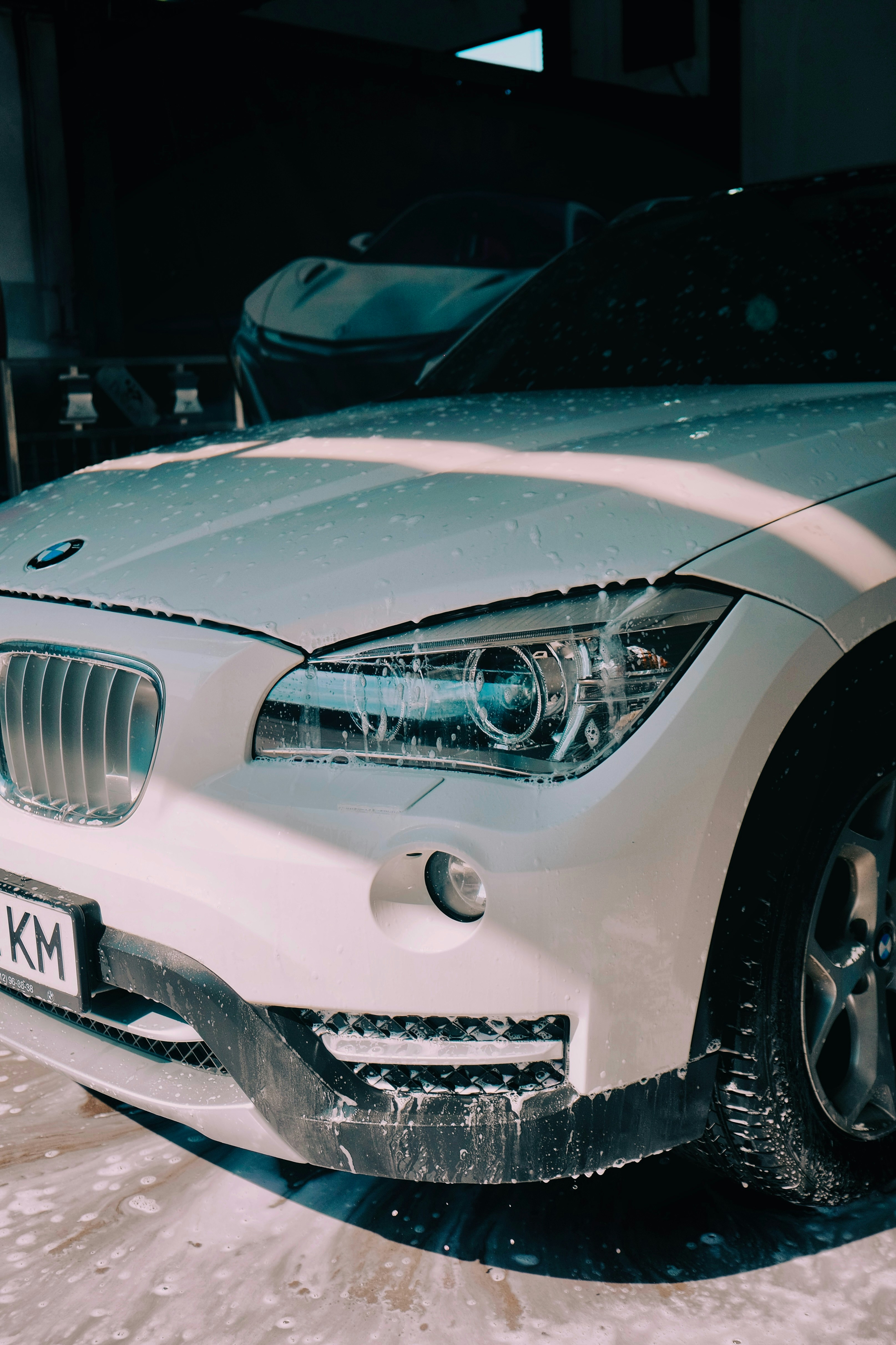 BMW car glistening with soap bubbles during a wash, showcasing its sleek lines and design. The sunlight casts intriguing shadows across the vehicle.