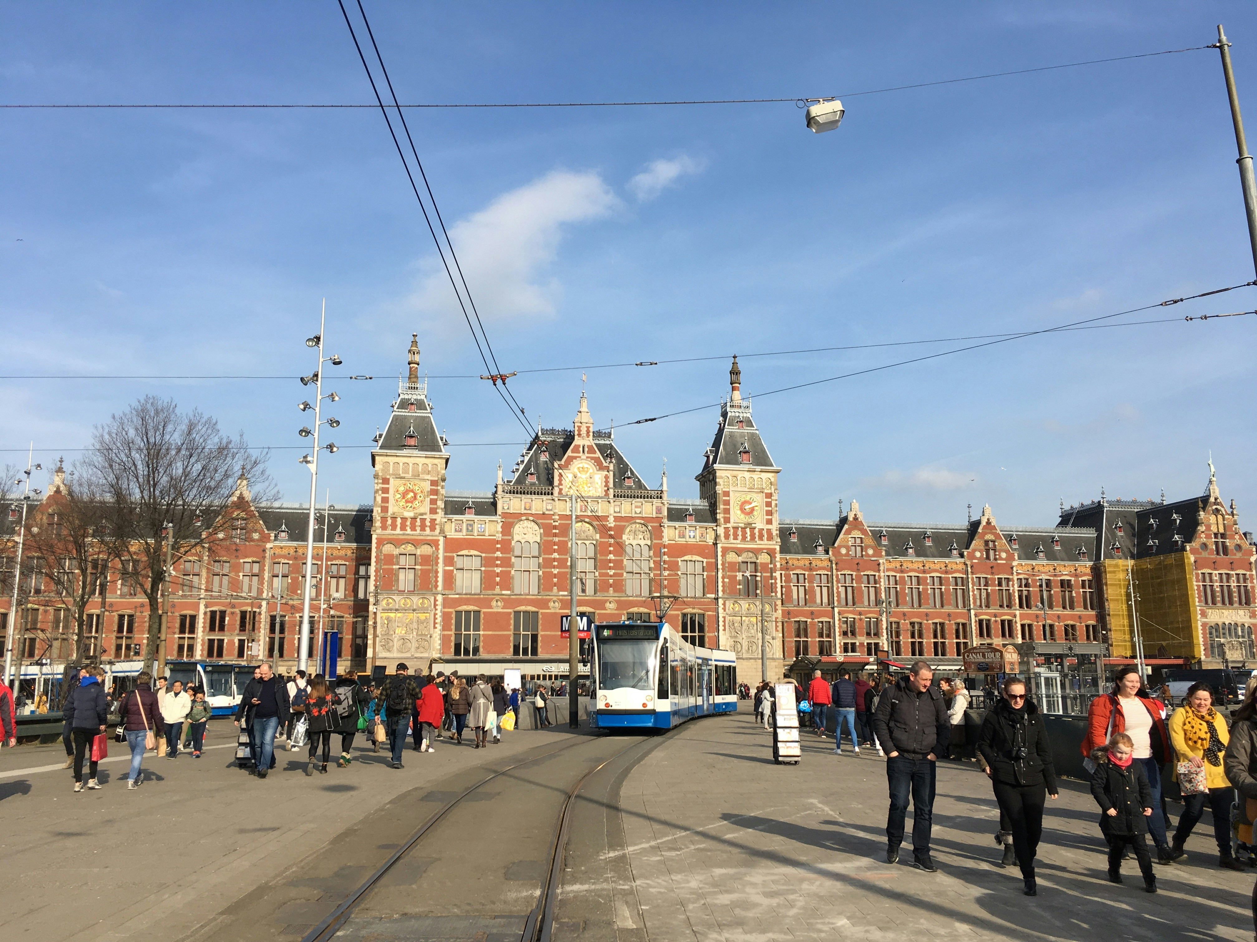 Busy city square with historic architecture, trams, and pedestrians under a clear blue sky.