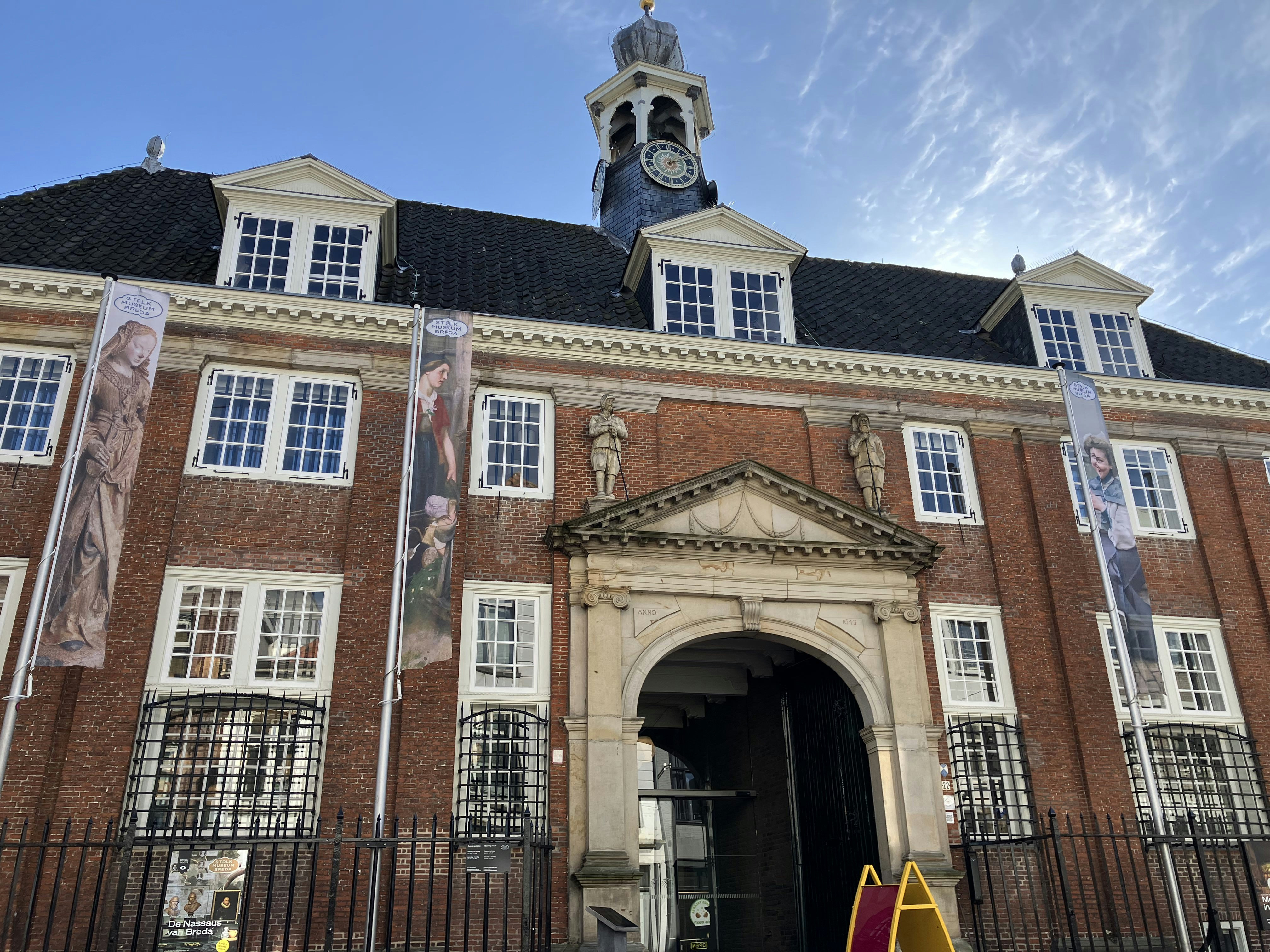 Grand brick building with a prominent clock tower and banners on either side, set against a blue sky.