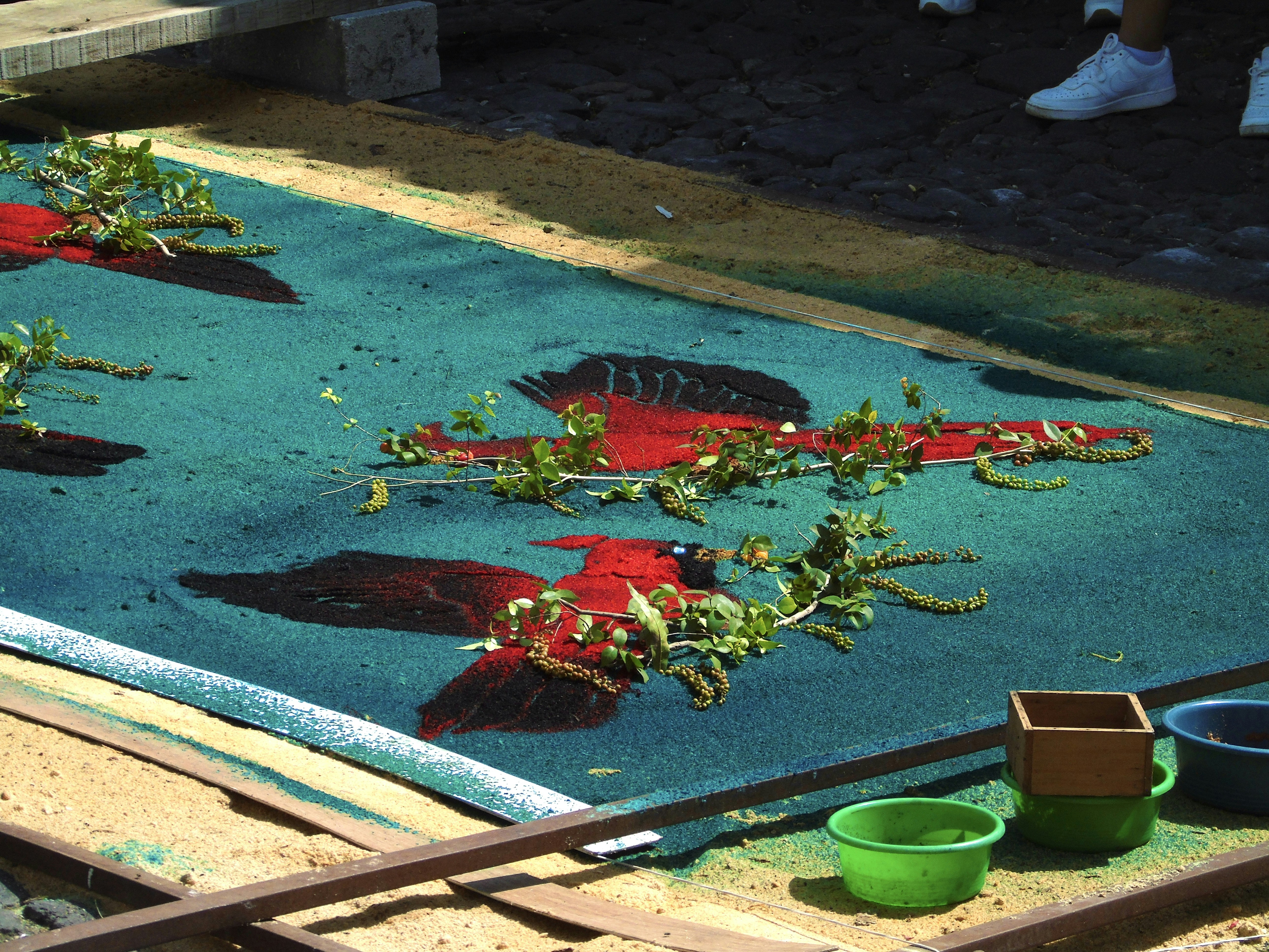 A photograph shows a teal mat with red shapes and scattered leaves, with green bowls and a small wooden box in the foreground and partial feet in sneakers in the background.
