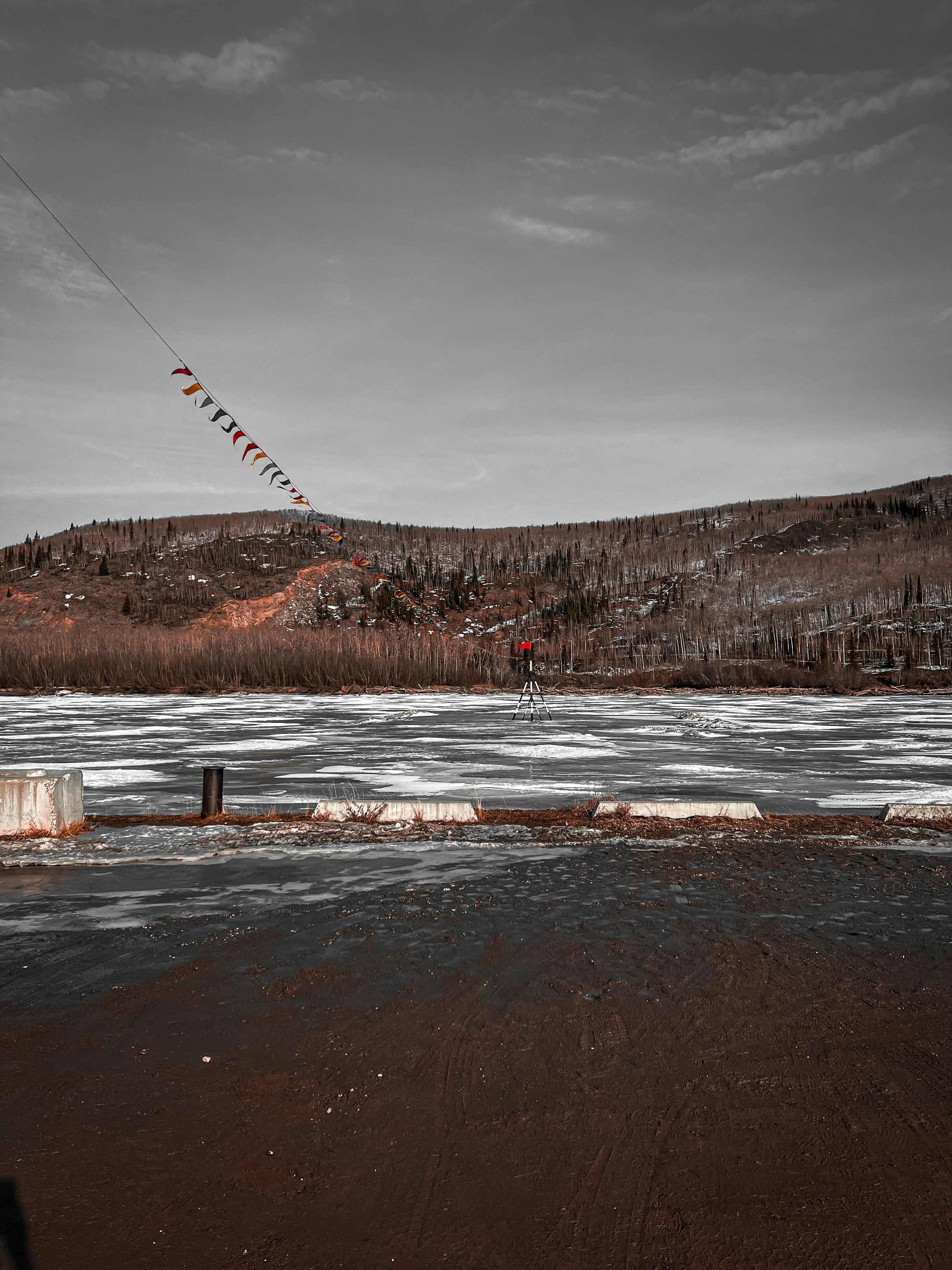 A lone figure stands on a frozen landscape, surrounded by muted colors and distant mountains, with colorful flags fluttering above. The stark contrast between the ice and the earthy tones evokes a sense of solitude.