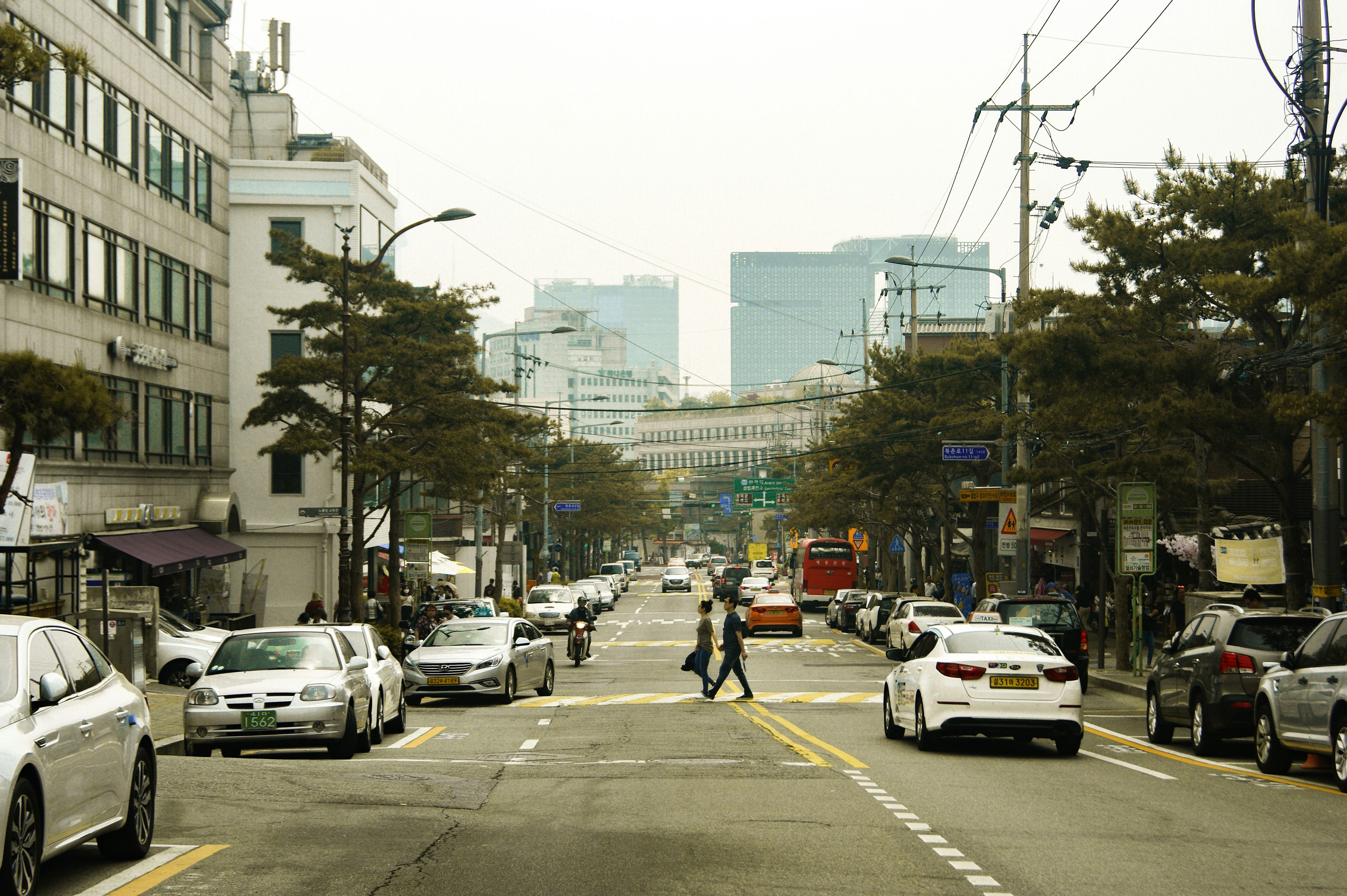 Urban street photograph taken on an overcast day, with parked cars lining both sides and two pedestrians crossing near the center.