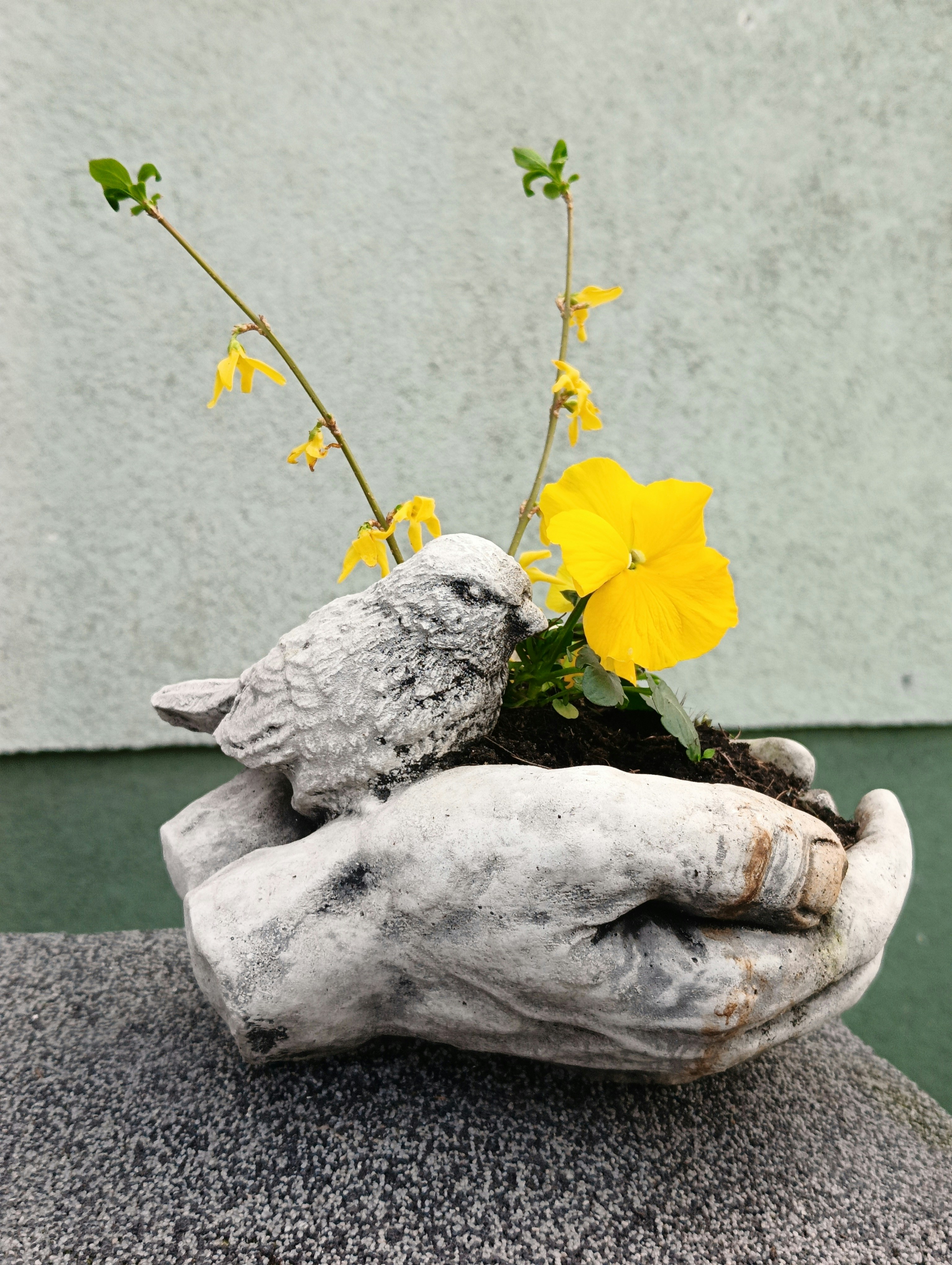 Weathered stone glove sculpture cradles bright yellow petunias, with a small bird perched atop, set against a pale wall.