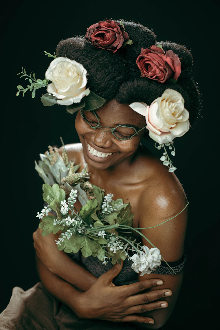Smiling woman wearing glasses and an elaborate crown of roses, holding a lush bouquet against a dark background.