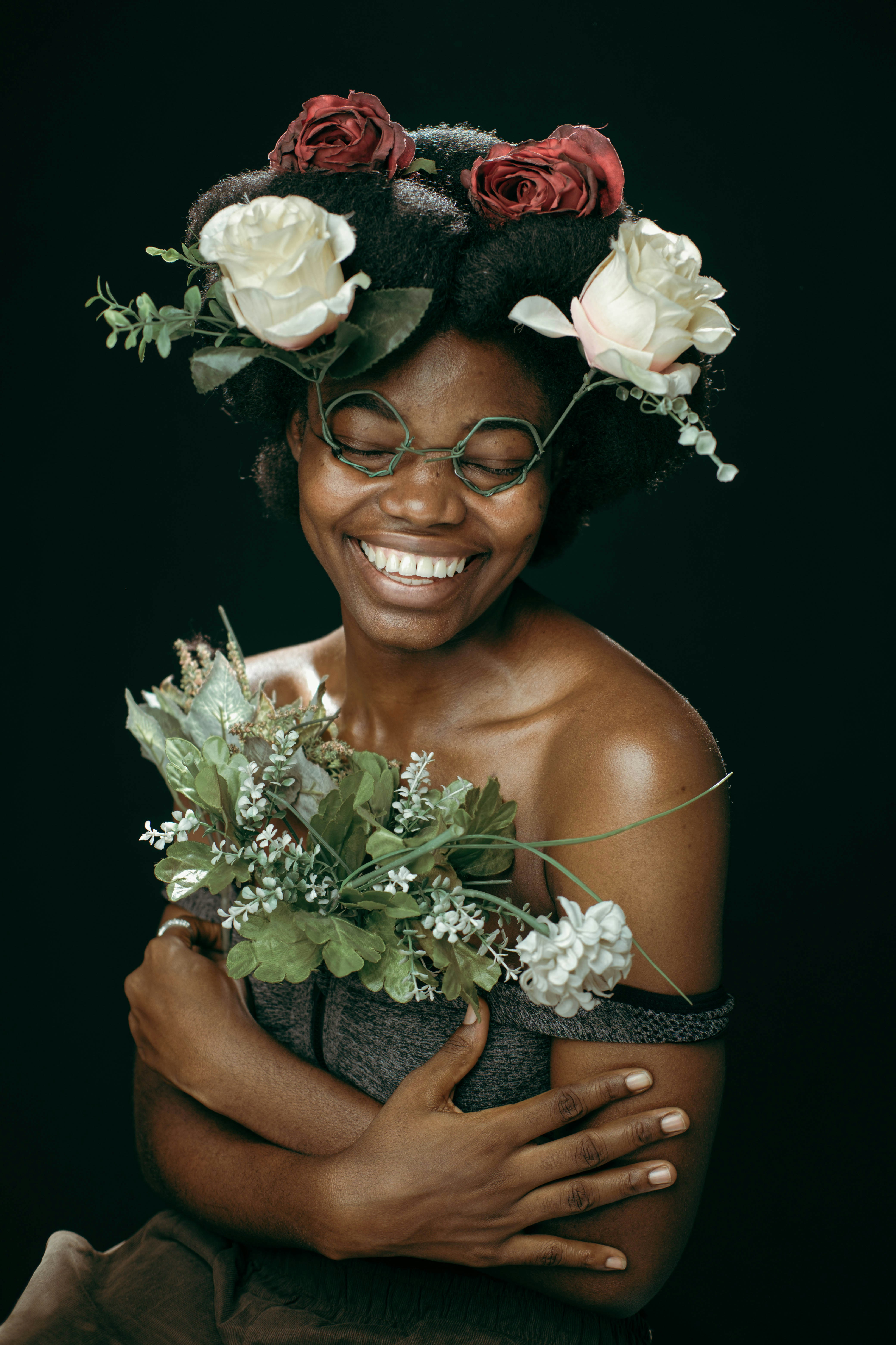 Woman with floral headpiece and greenery, smiling warmly against a dark background.