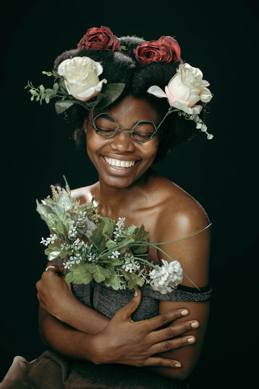 Smiling woman with closed eyes wearing round glasses and a crown of red and white roses, holding a bouquet against a dark background.