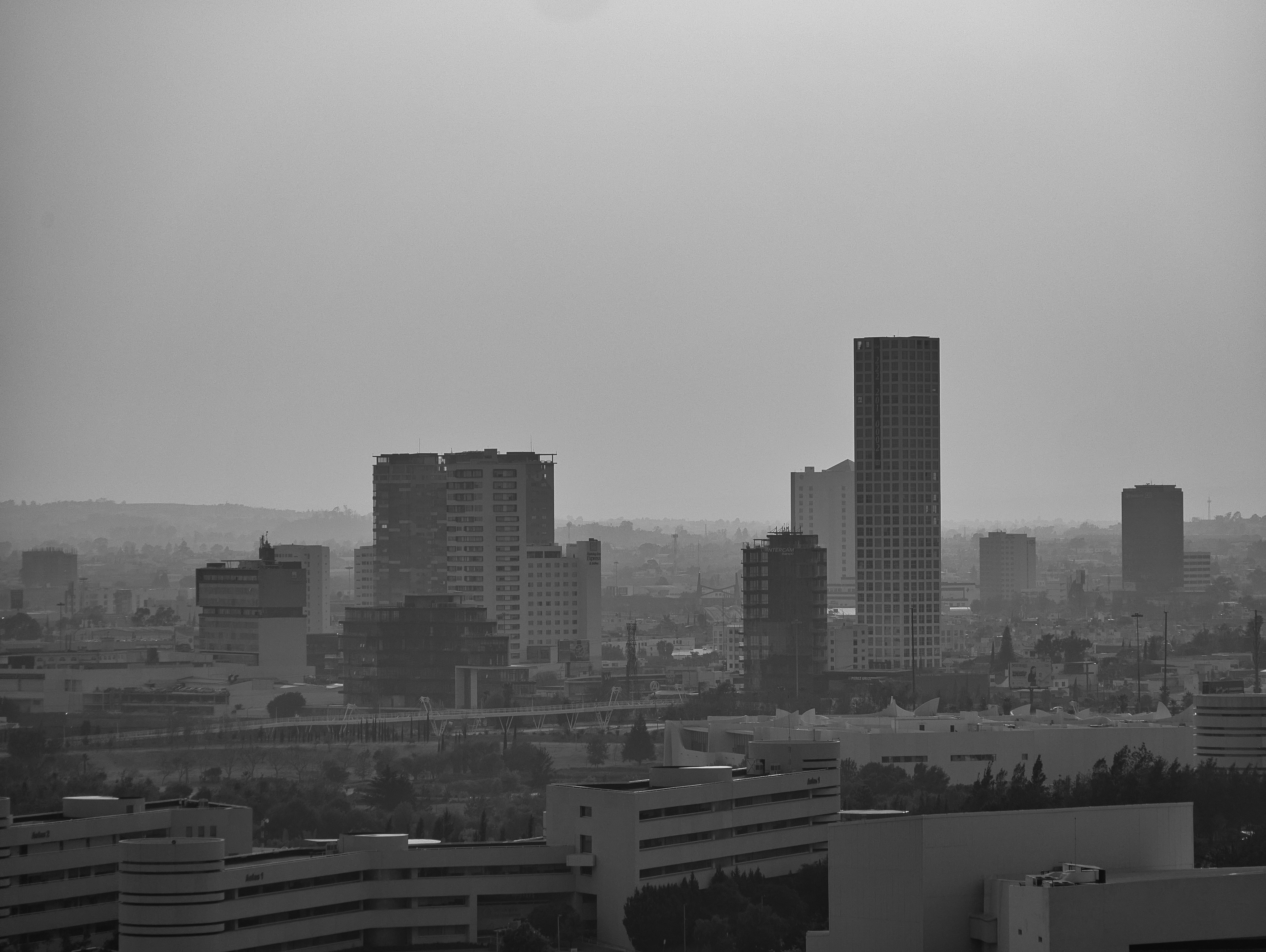 Grayscale cityscape photograph featuring a cluster of high-rise buildings with a dominant slender tower piercing the hazy skyline. The composition emphasizes geometric silhouettes against a muted backdrop.