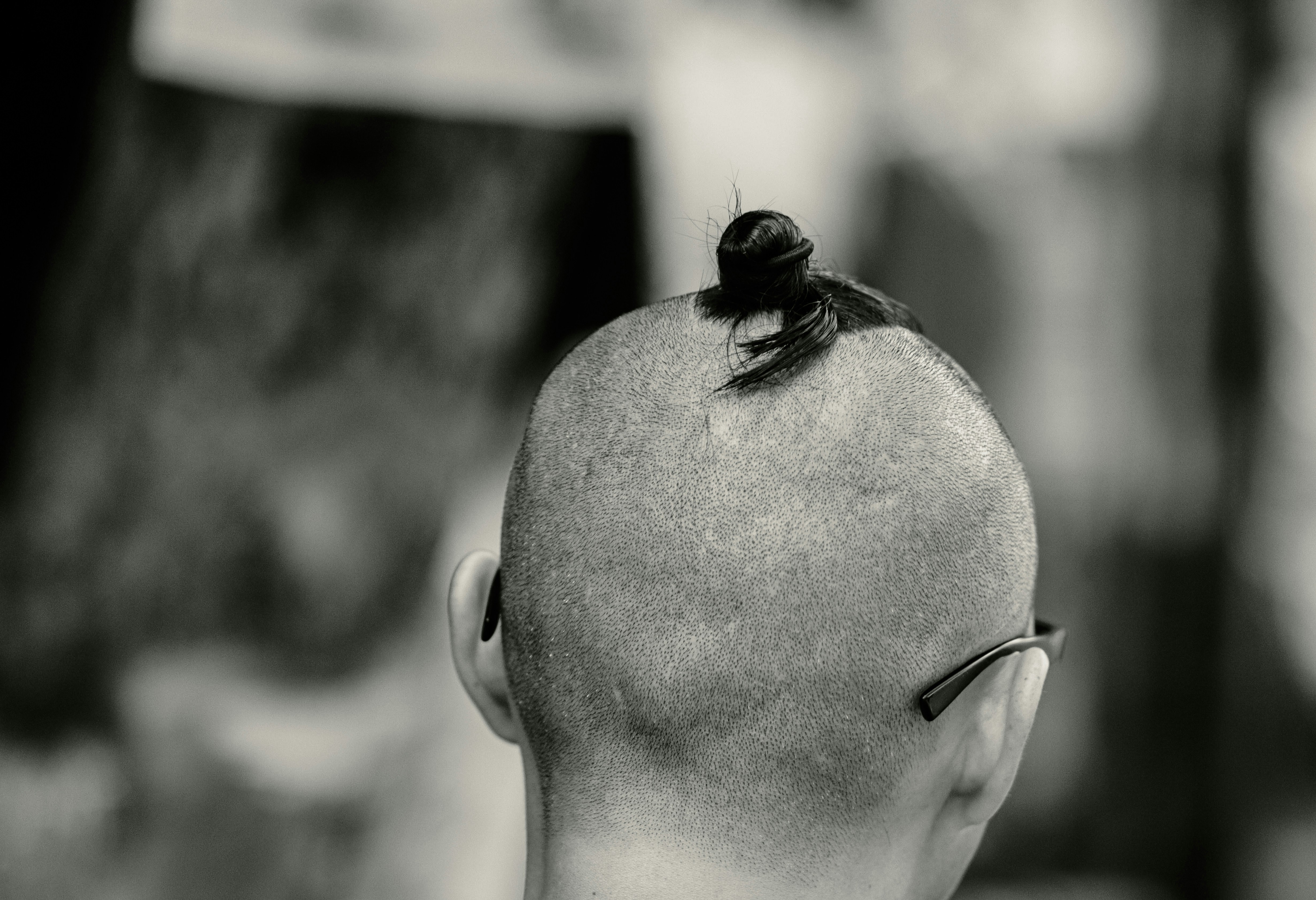 Close-up of a shaved head with a small topknot, showcasing a unique hairstyle that blends contemporary and traditional elements. The monochrome tones enhance the texture and details of the scalp.