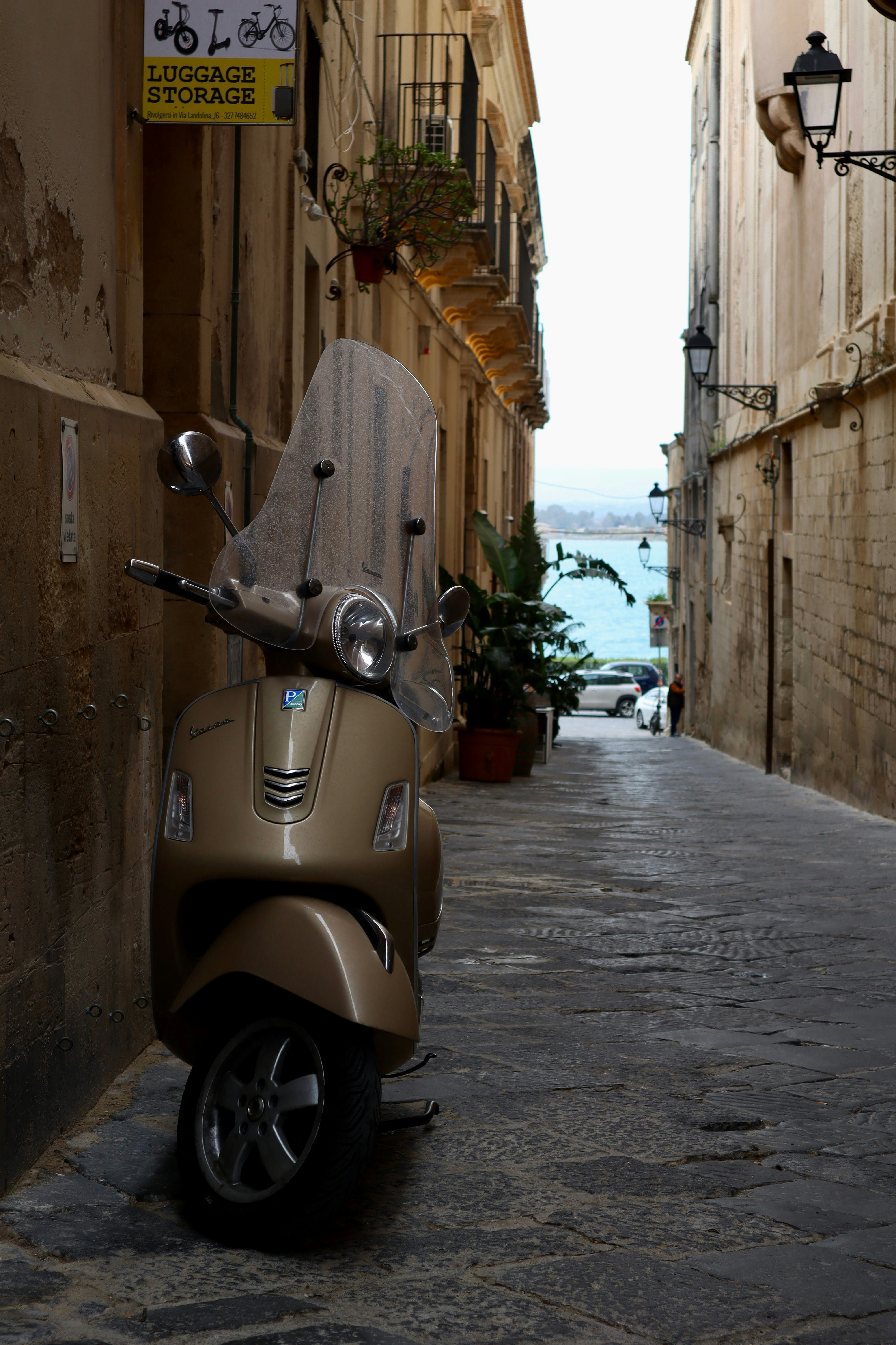 A vintage scooter parked in a narrow, sunlit alleyway lined with stone walls and potted plants, leading to a glimpse of the sea.