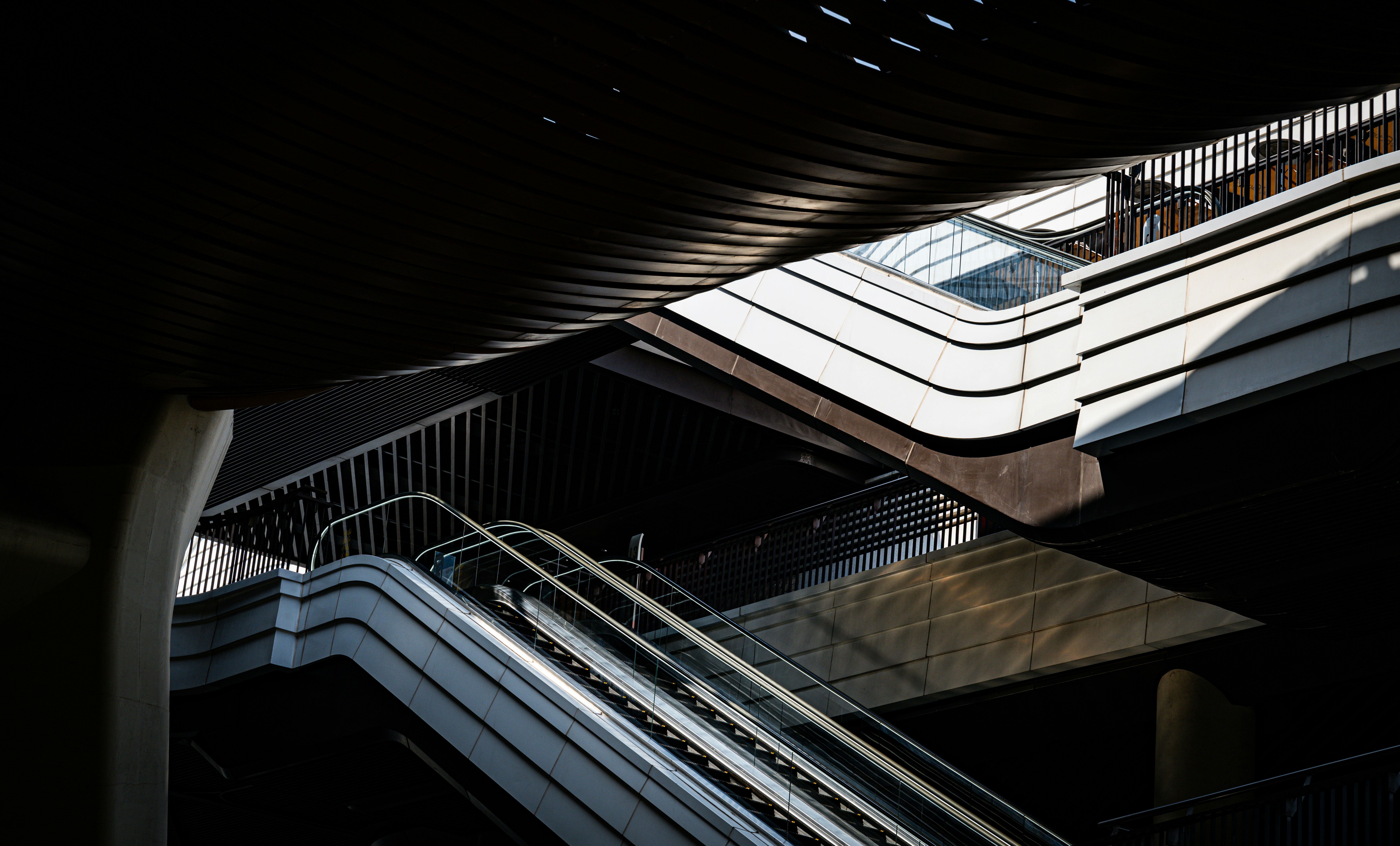 This striking image captures the intricate interplay of shadows and light on a modern architectural structure, featuring sleek escalators and curved lines. The composition highlights the contrast between the dark, shadowy areas and the bright, sunlit surfaces, creating a dynamic and visually engaging scene. The use of muted colors and sharp angles enhances the modern and futuristic atmosphere of the setting, making it a captivating piece of urban photography.
