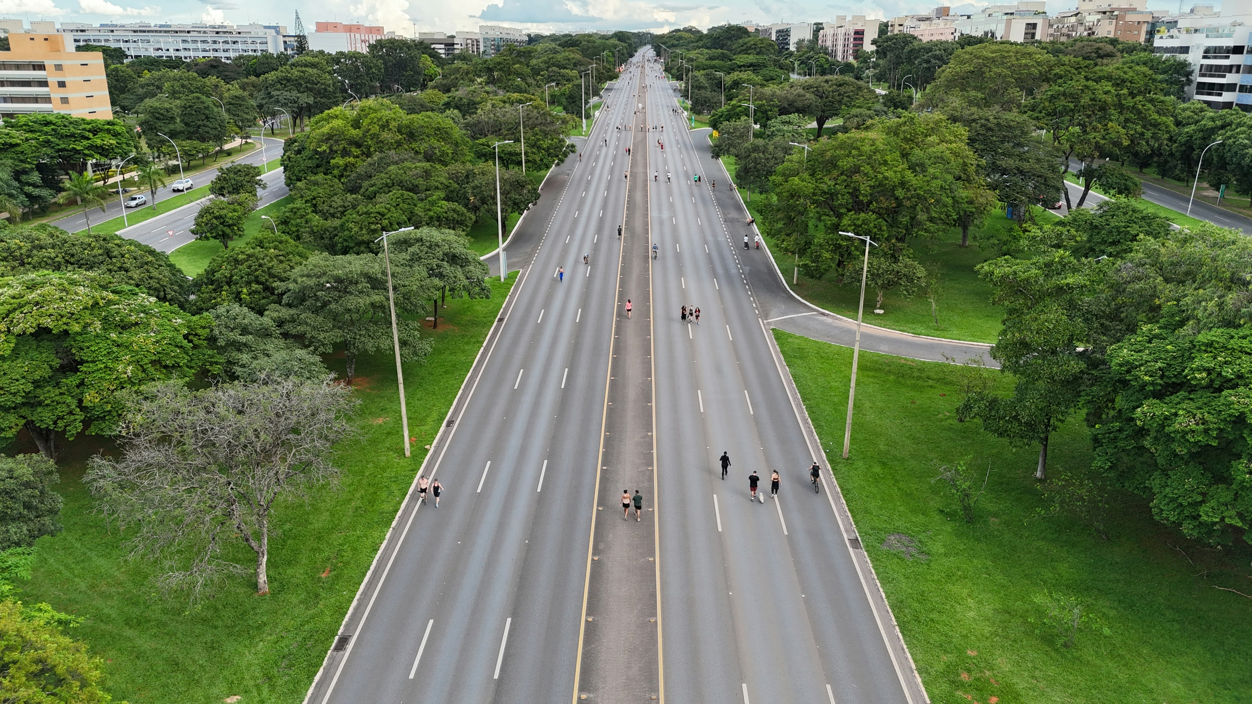 Aerial view of a wide, empty roadway flanked by lush green trees and urban architecture under a cloudy sky.