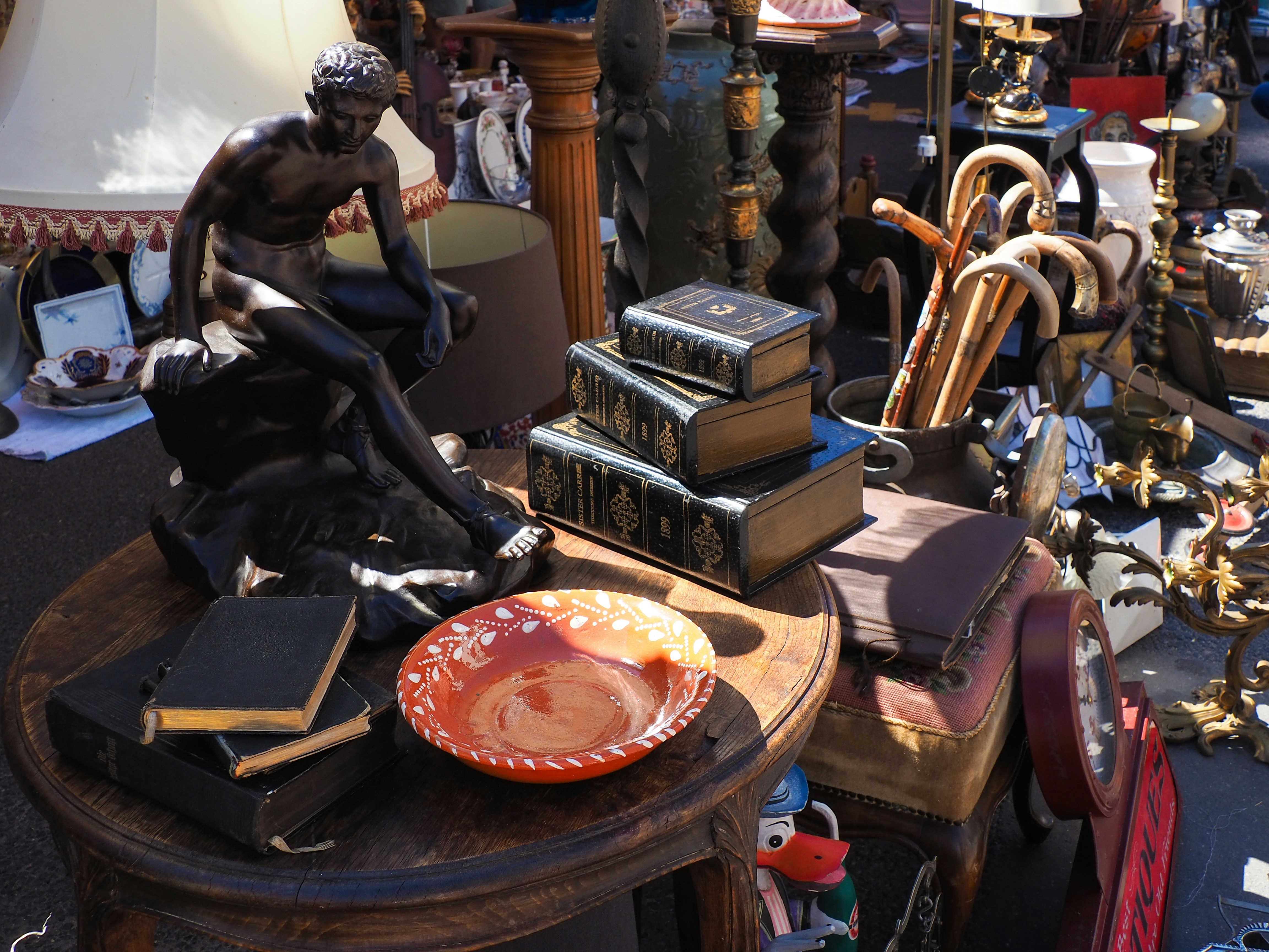 Bronze Thinker statue sits on a wooden table among stacked books, a red plate, and assorted antique curios at a bustling market stall.