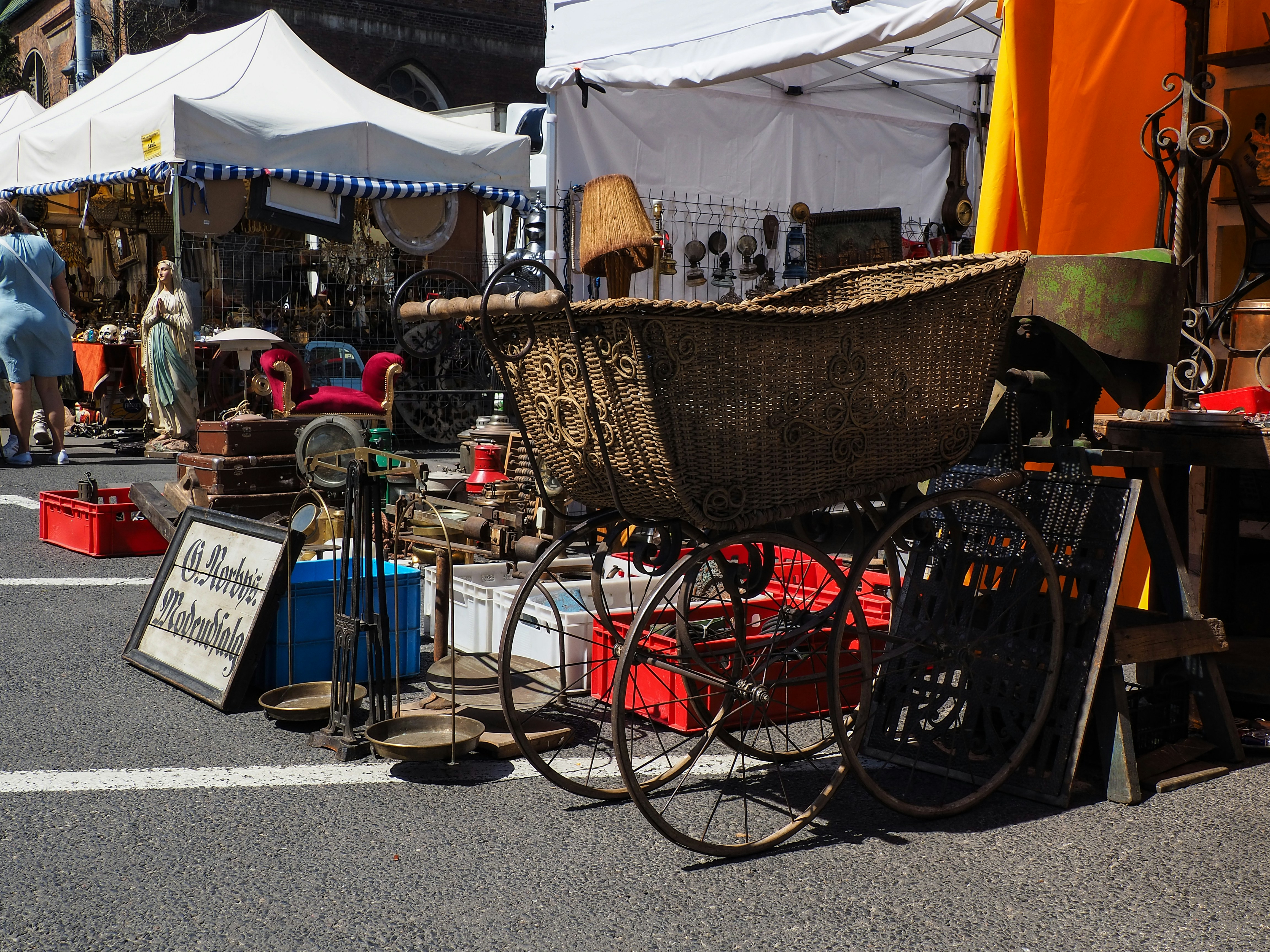 Busy outdoor market scene with white canopies, vintage wicker carts, and assorted goods lining a sunlit street.