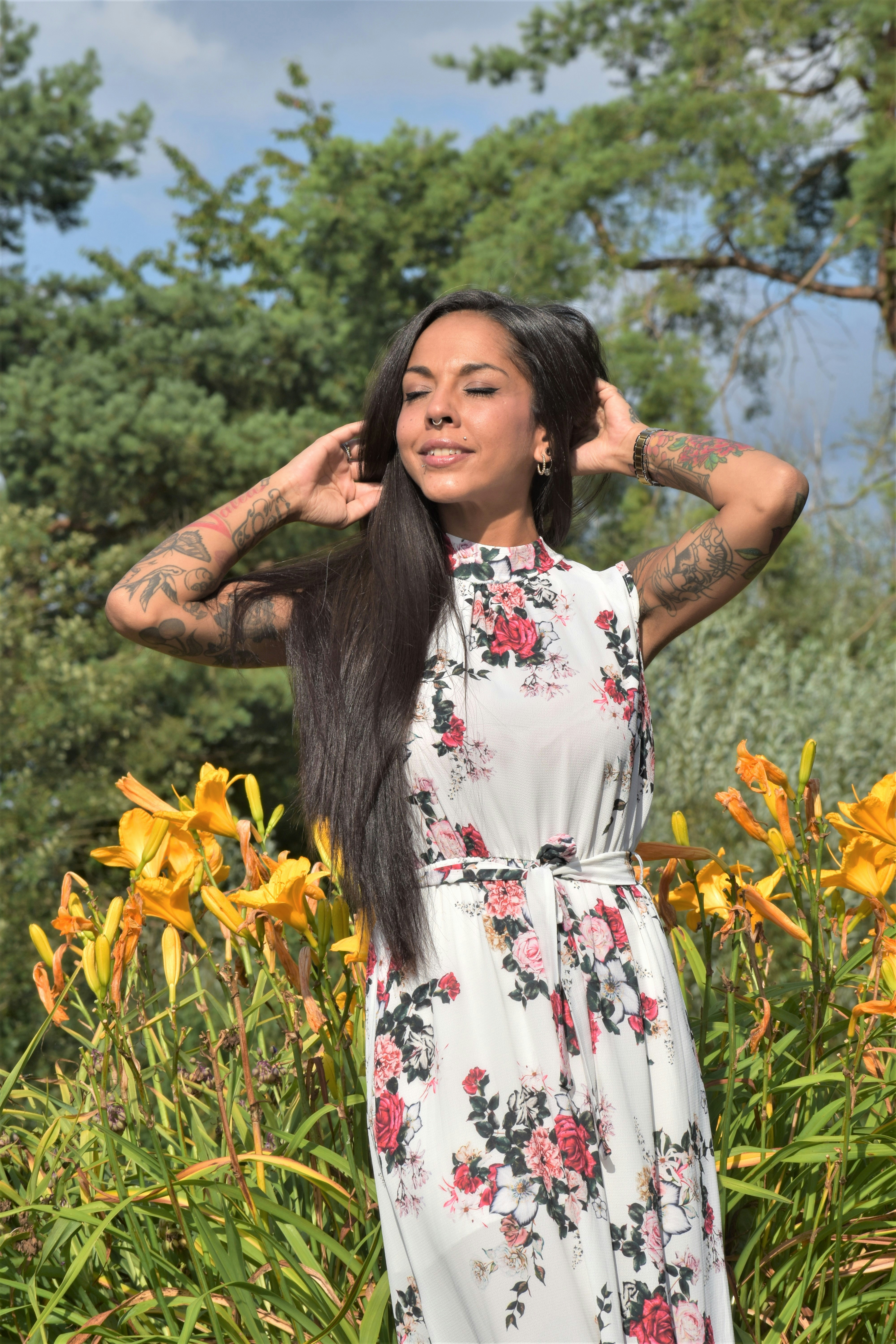 Portrait of a tattooed woman in a white floral dress standing among tall yellow lilies under bright daylight. The composition emphasizes natural garden surroundings and the subject's relaxed, confident pose.